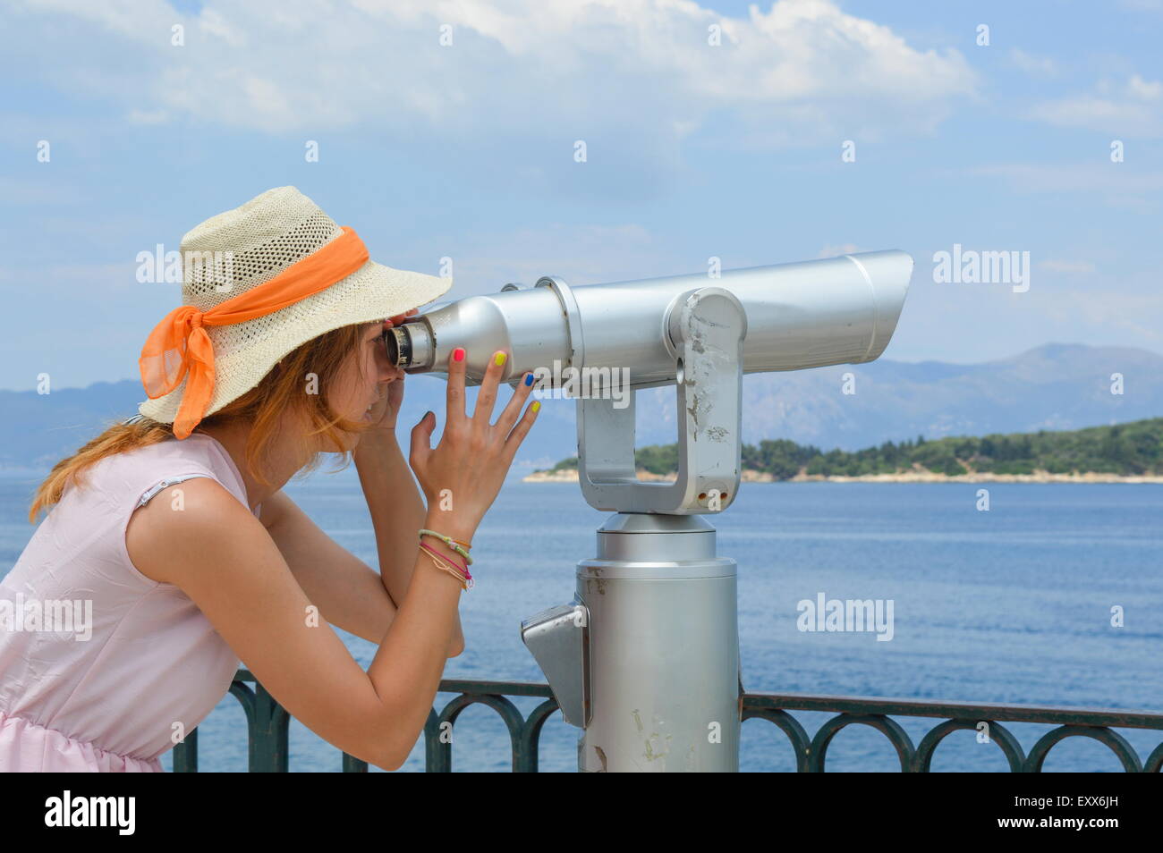 Young girl looking thru public binoculars at the seaside wearing straw ...