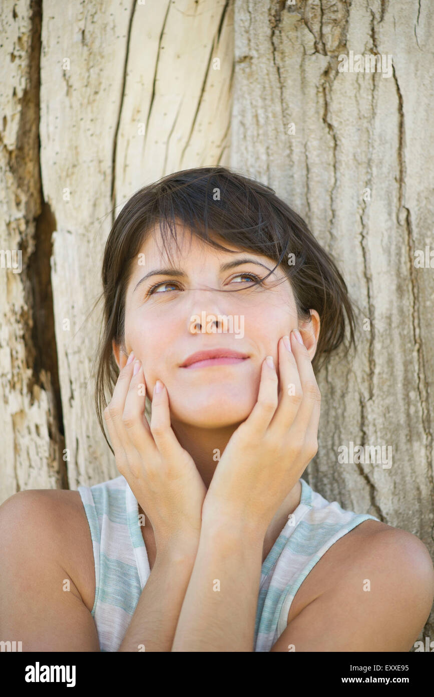 Woman leaning against tree trunk with face in hands, looking up ...