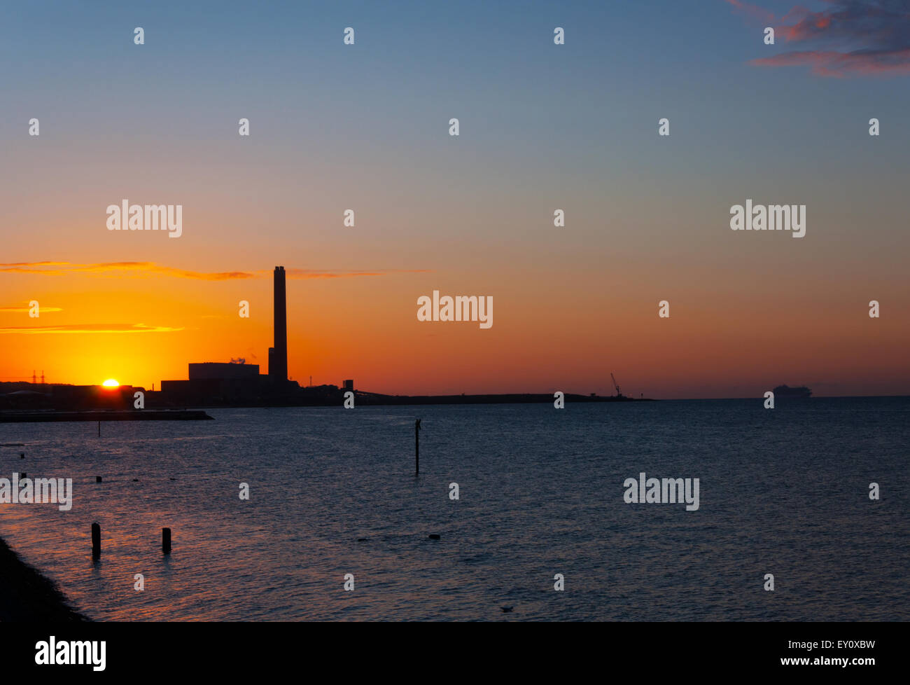 Sunrise behind a factory at the seaside with far cranes and a ship ...