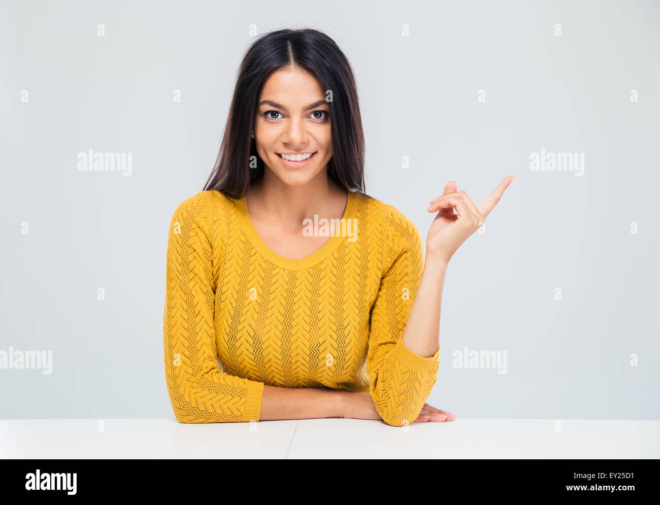 Smiling young woman sitting at the table and pointing finger away over ...