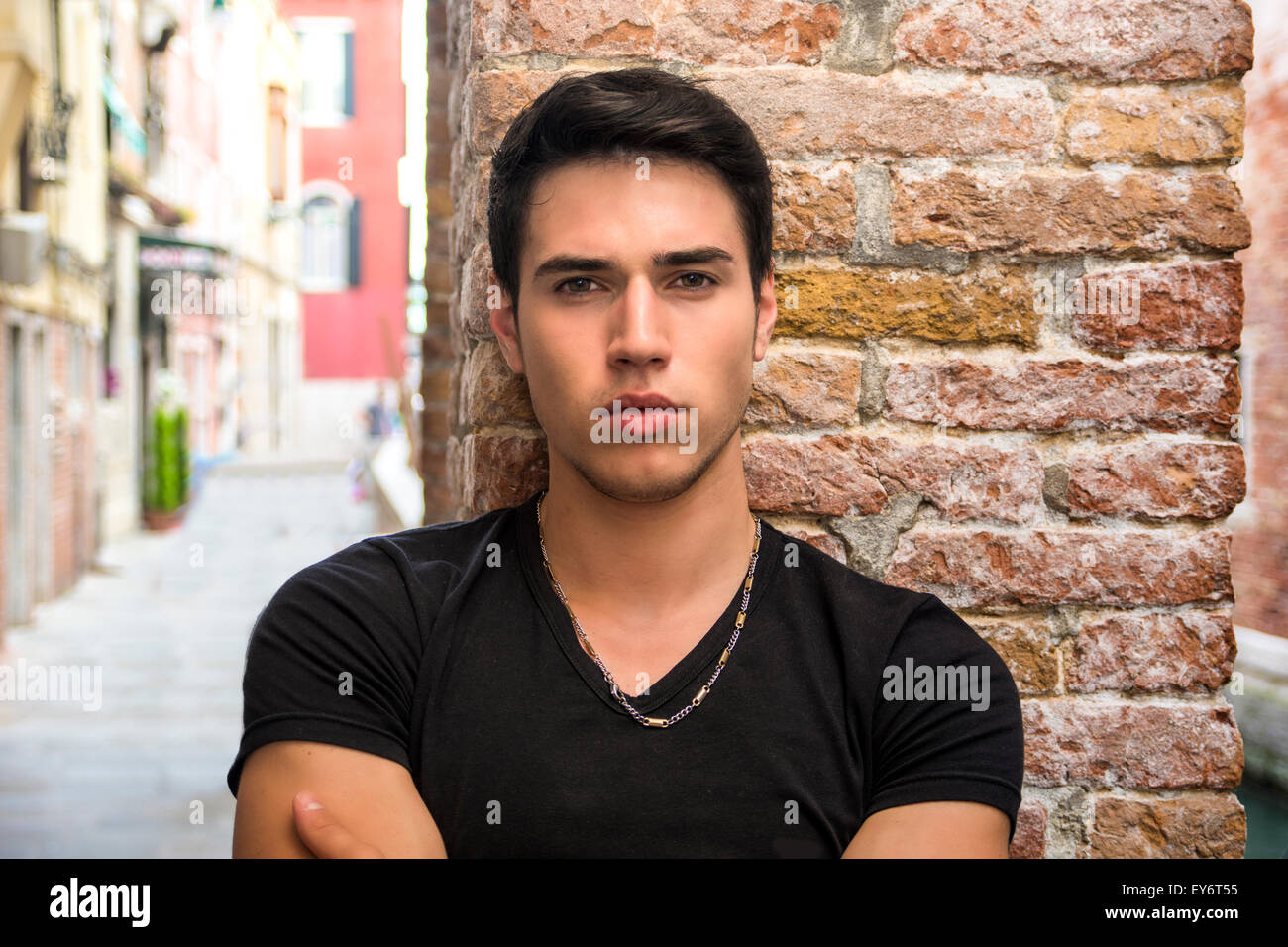 Attractive young man sitting against brick wall, looking at camera ...