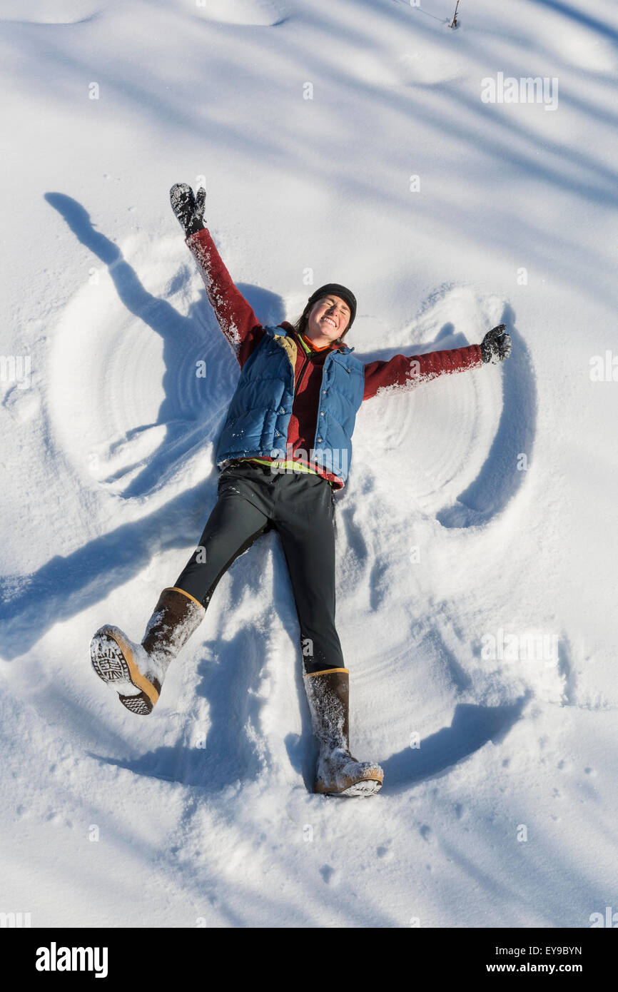 A smiling young woman wearing a hat and gloves makes a snow angel in a ...