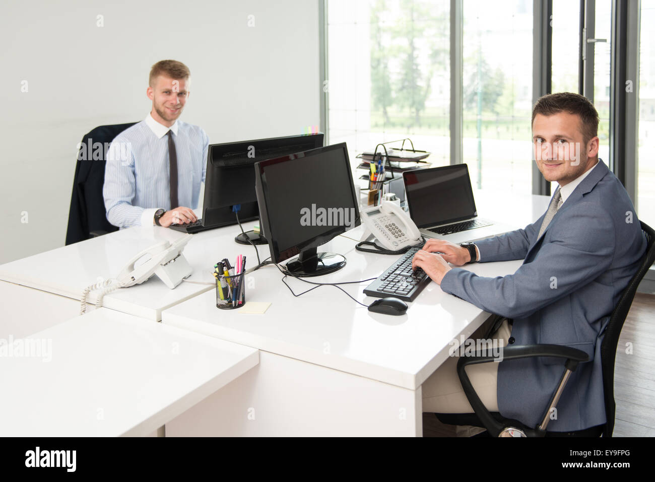 Two Businessmen In An Office Smiling At The Camera While Working ...