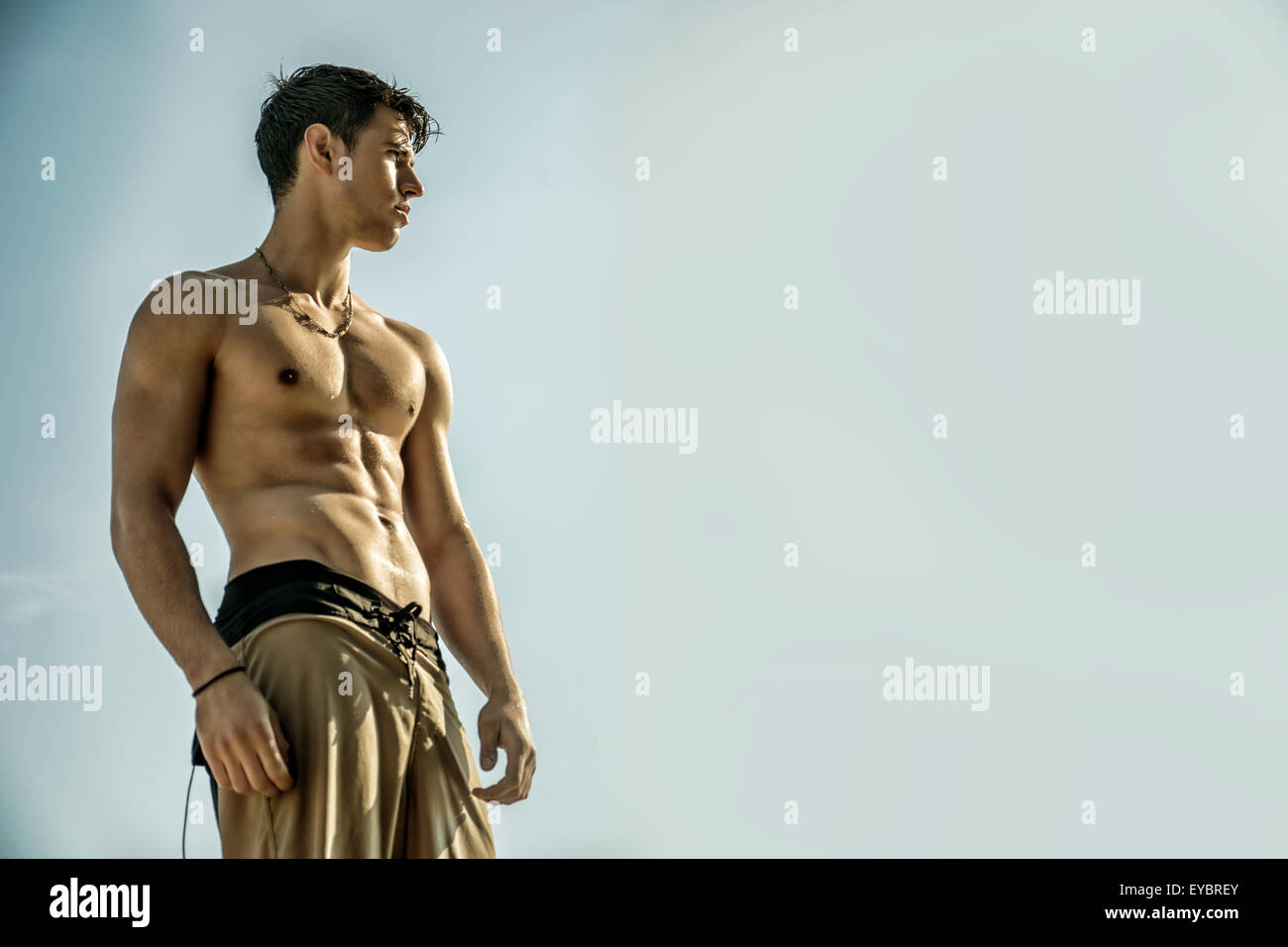 Muscular young man shirtless standing against blue sky, seen from below ...