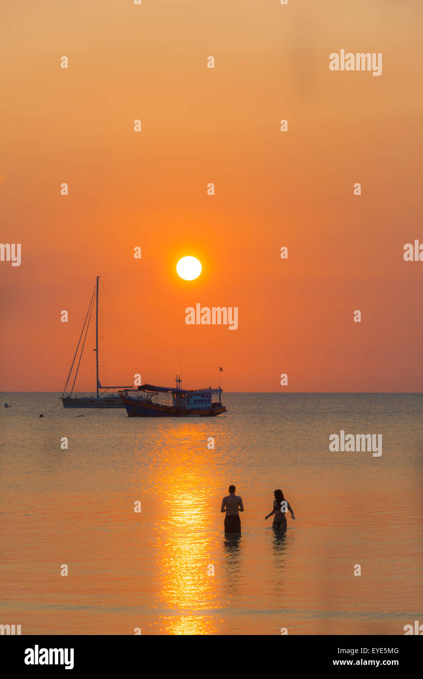 Two people standing in the water, boats in the sea at sunset, Koh Tao ...
