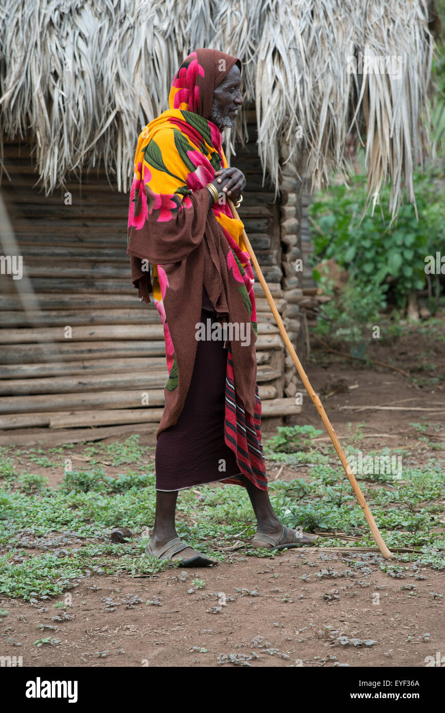 Datoga elder in brightly coloured robe leans on cane in front of hut ...