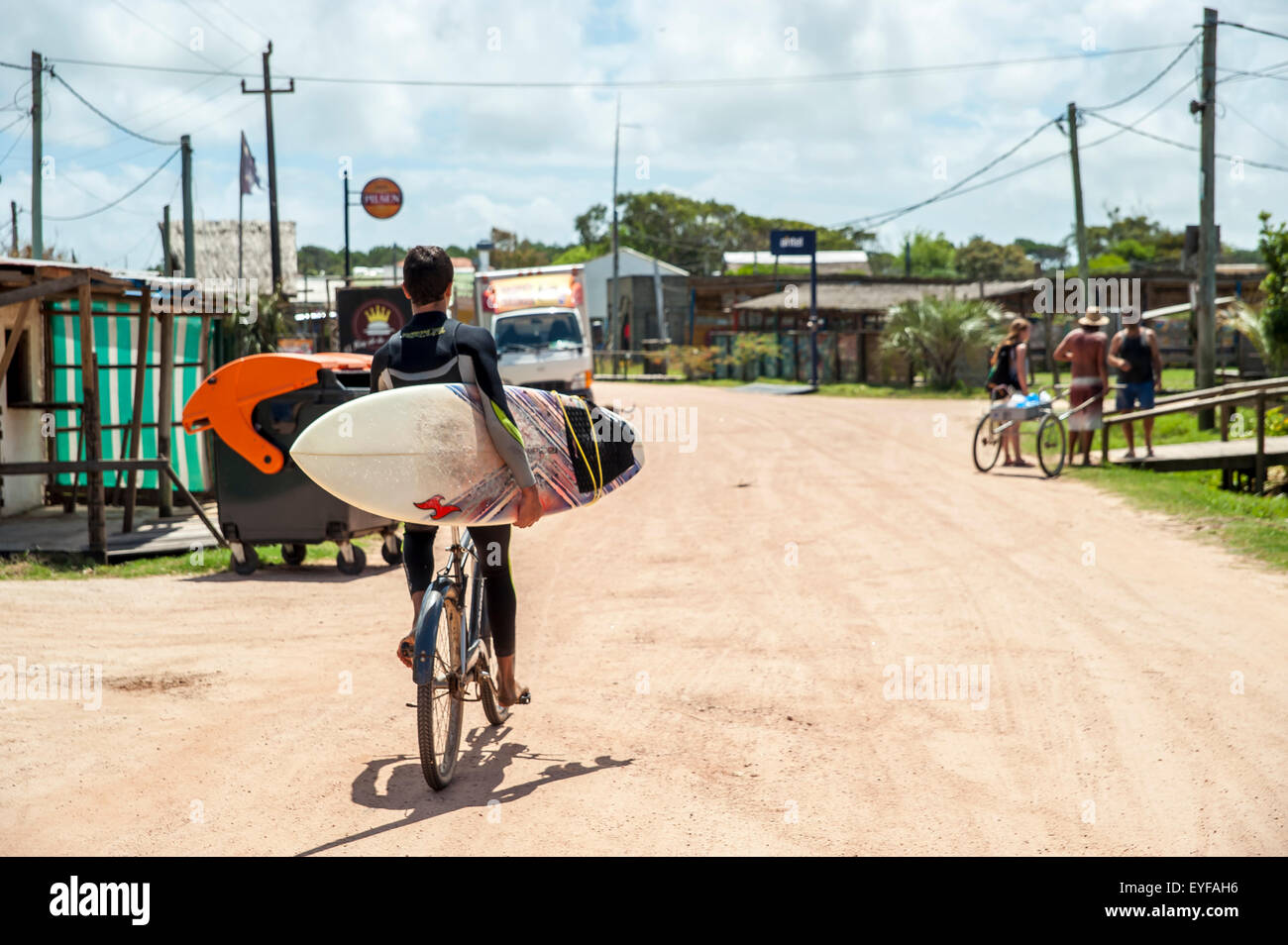 A surfer cycles down a dirt road on his bicycle holding his surfboard ...