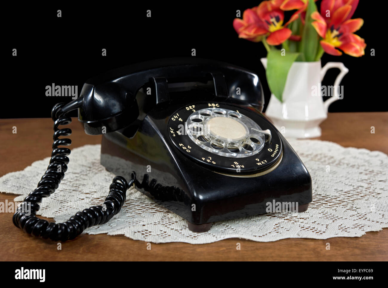 Black rotary dial telephone on lace doily with tulip bouquet in pitcher ...