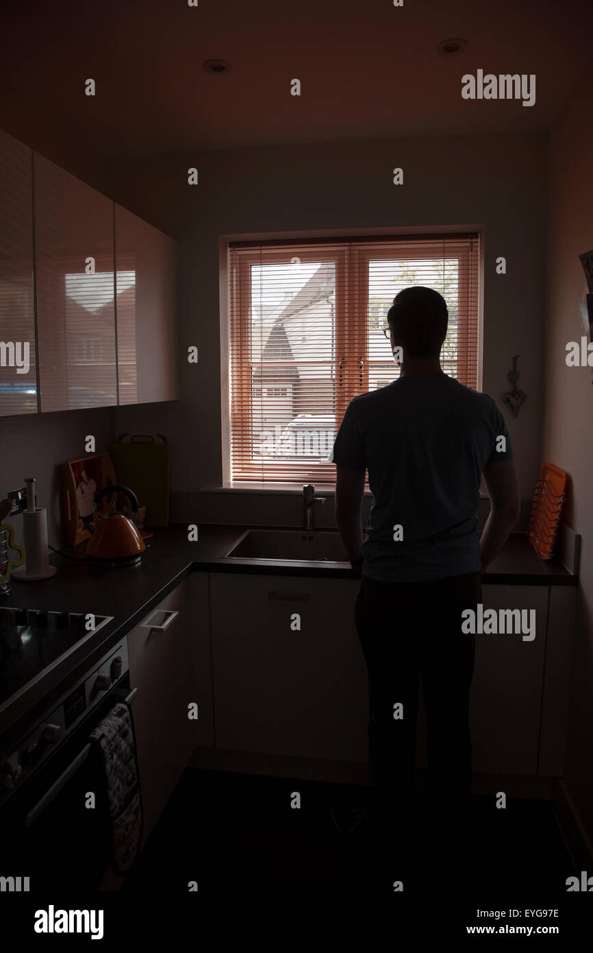 Young man in a kitchen looking out through a window blind. Portrait ...