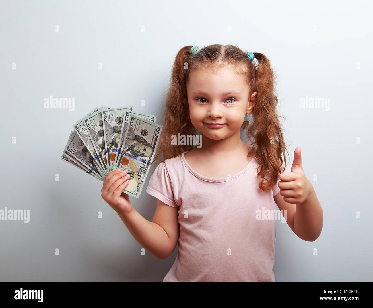 Happy rich kid girl holding money and showing thumb up sign on blue ...