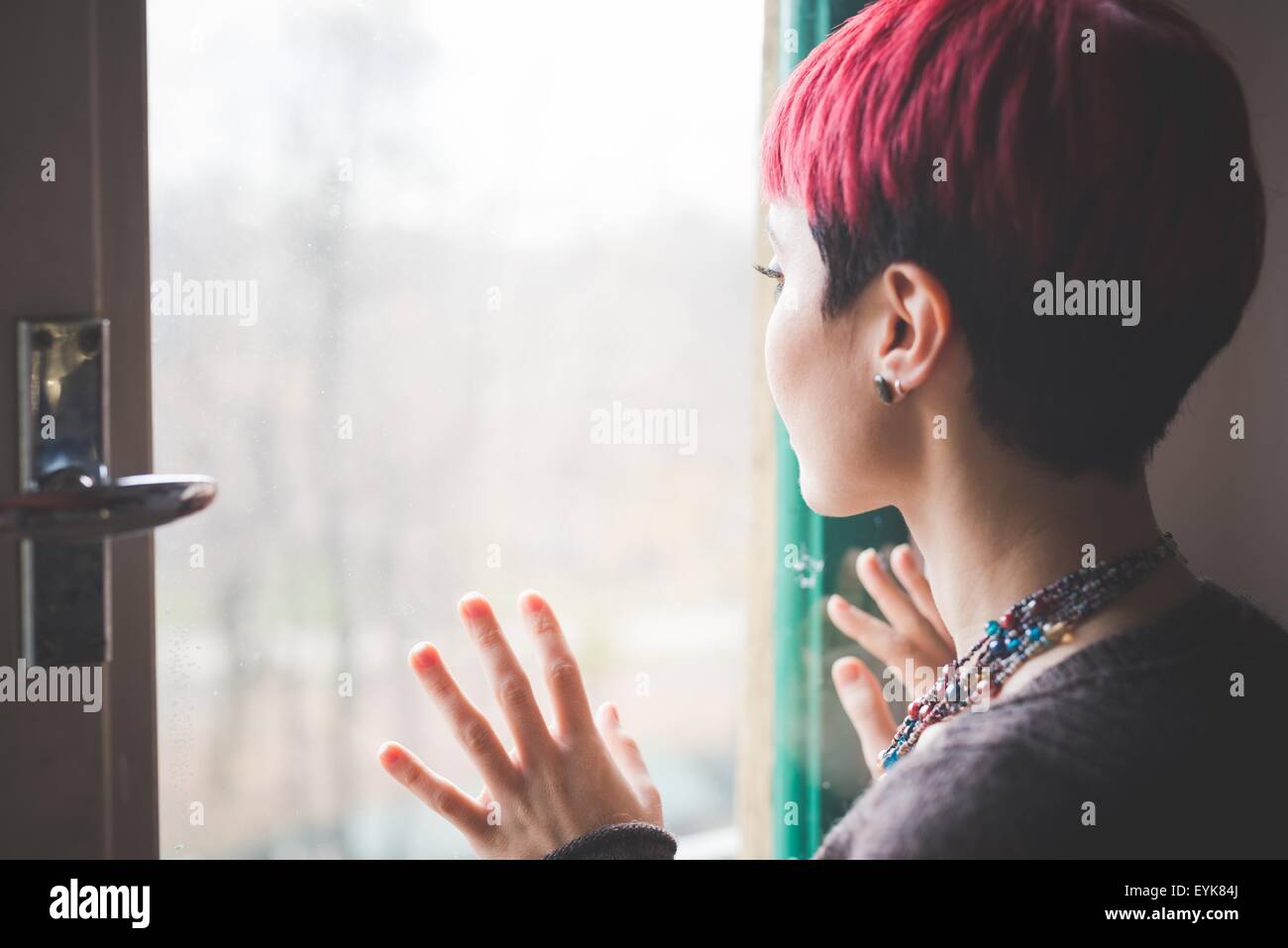 Young woman looking out of window, hands touching window, rear view ...