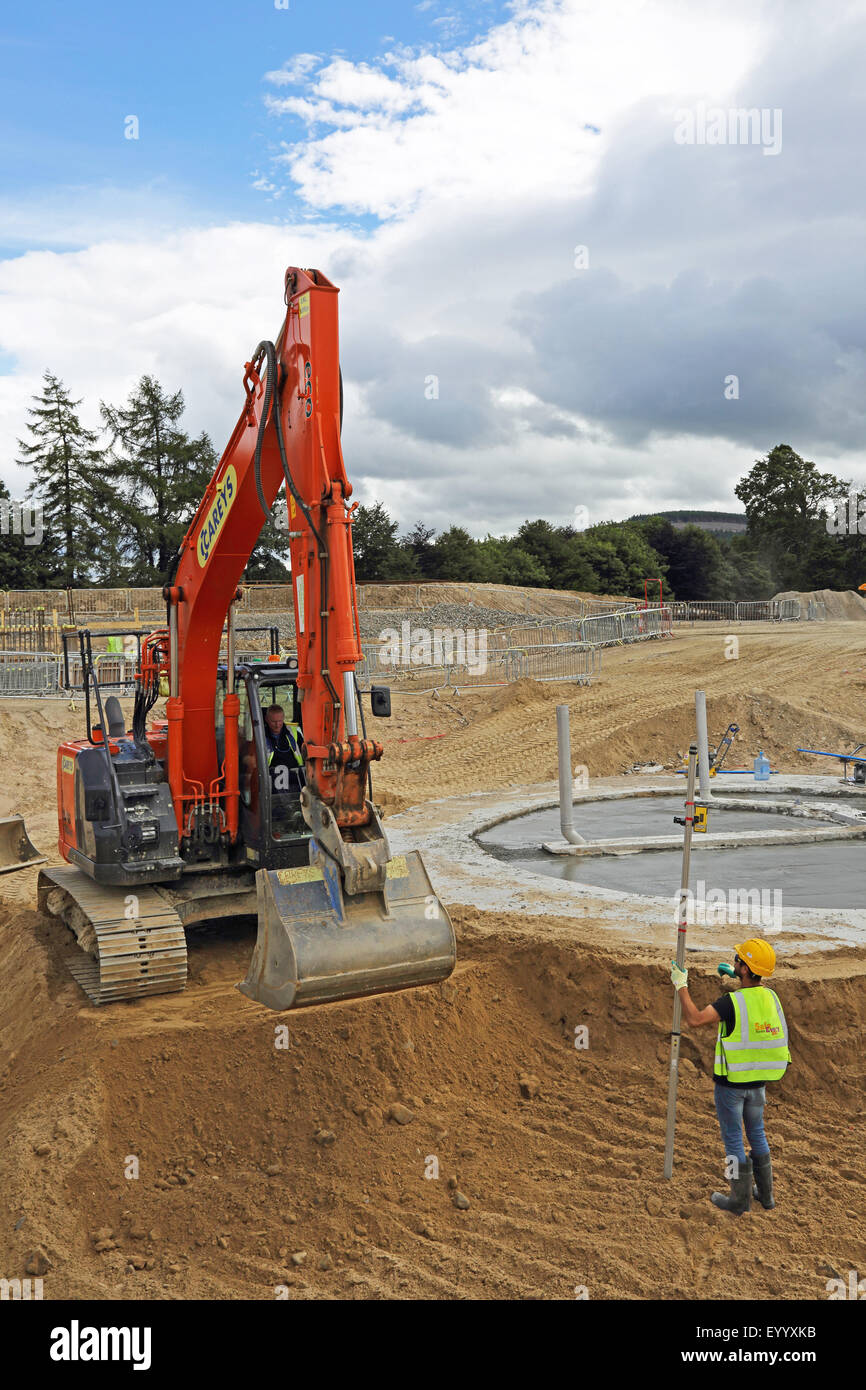 A surveyor holds a ranging pole in front of a large excavator on an ...