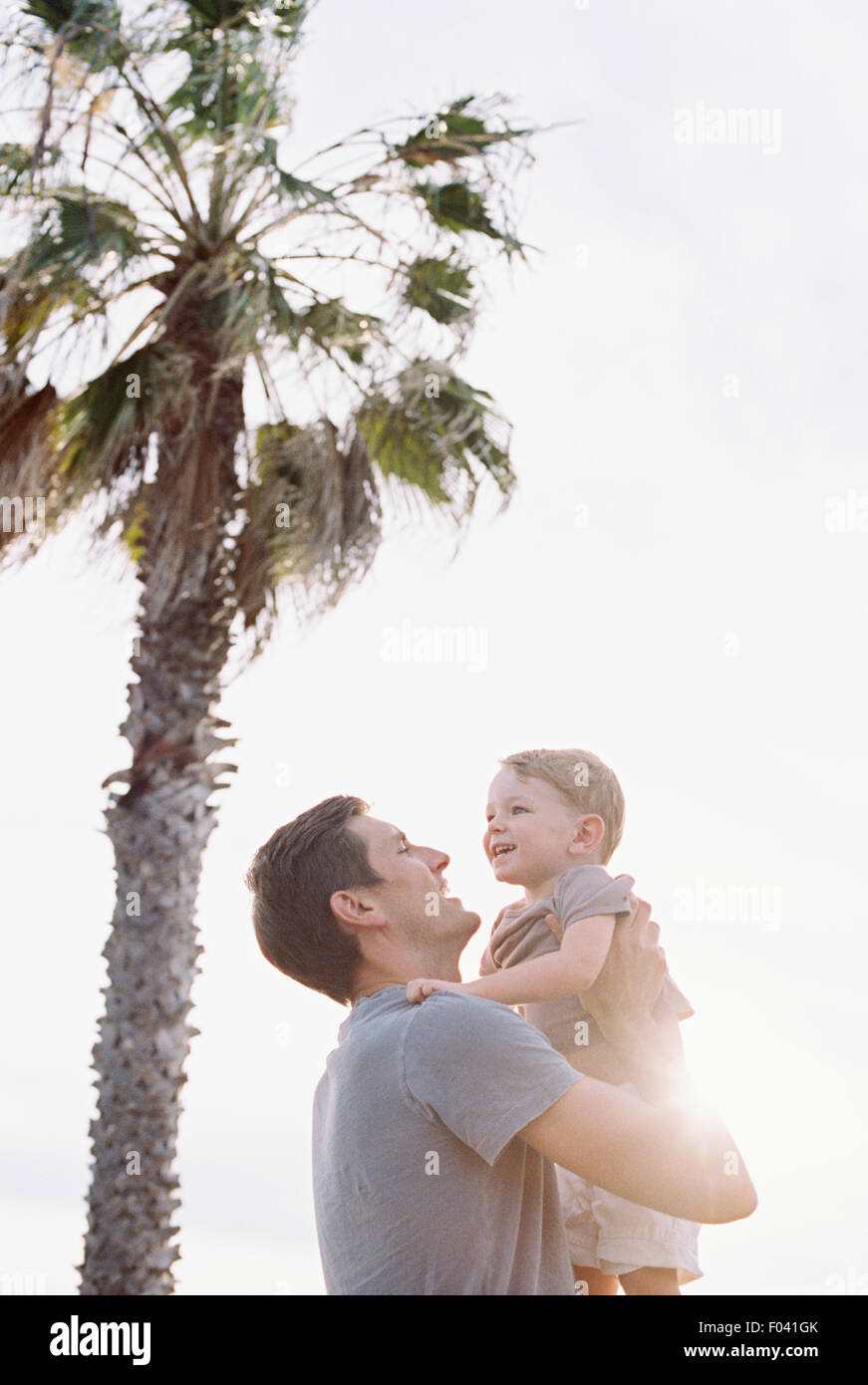 Smiling man standing by a palm tree, carrying his young son in his arms ...