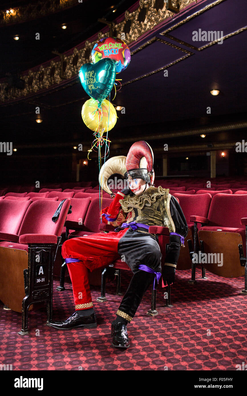 A sad jester sits in a theatre holding balloons that say 'Welcome Back ...