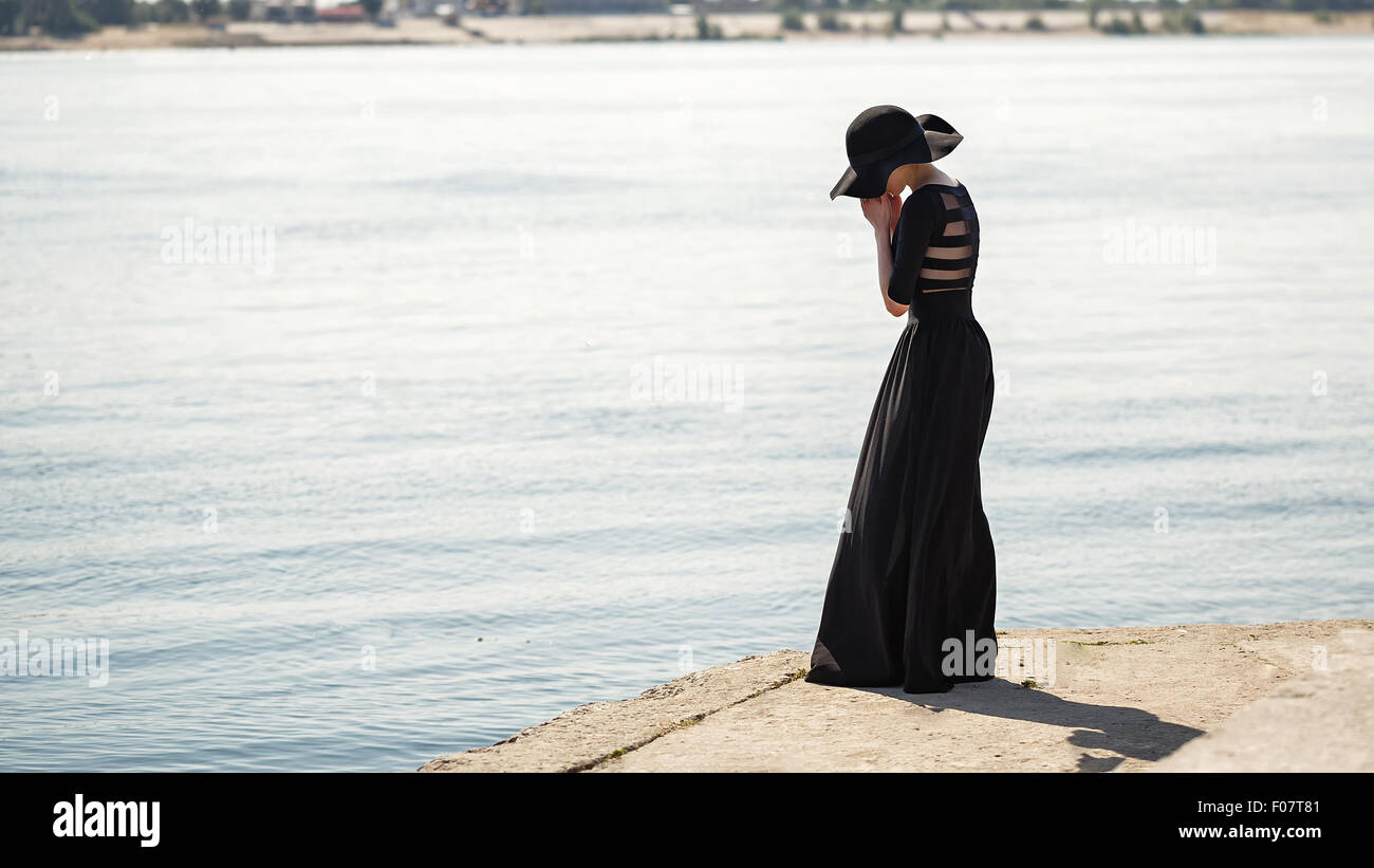 Young slender dancer in a hat and a long black dress sad on the ...