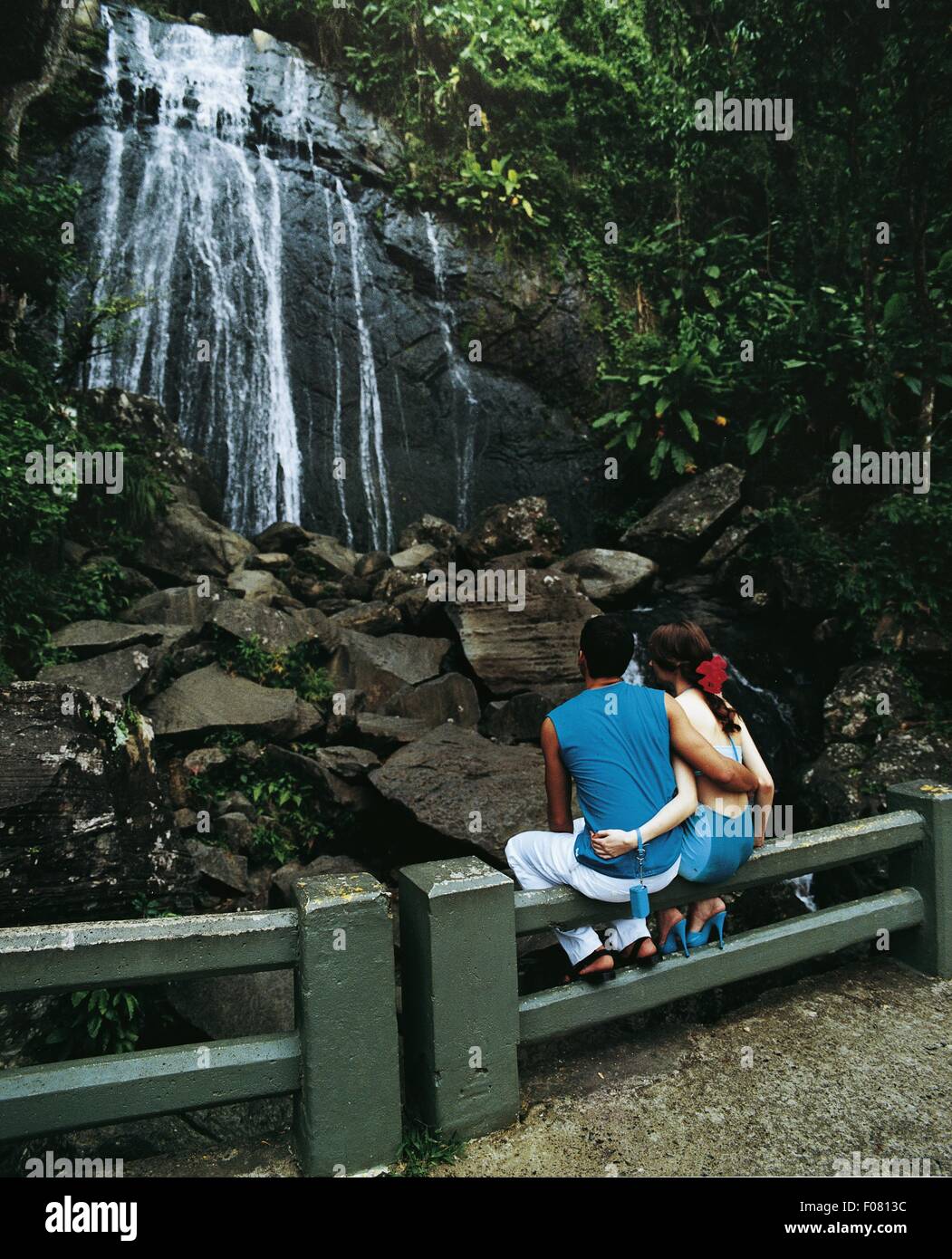 Couple sitting on fence in front of waterfall Stock Photo - Alamy