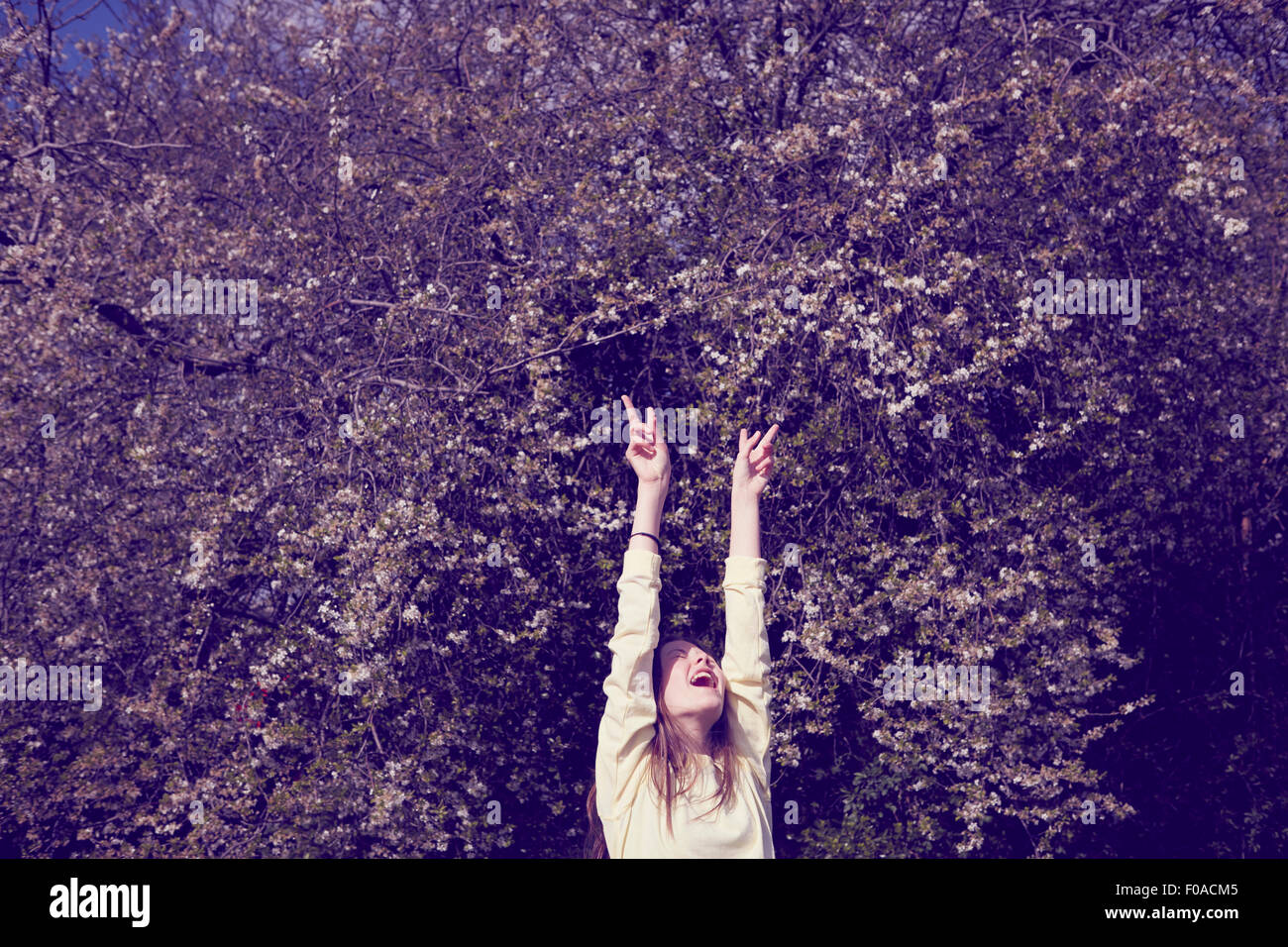 Portrait of girl in front of foliage with arms raised making peace sign ...