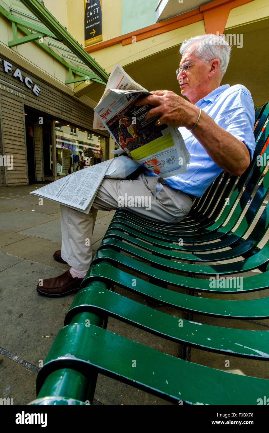 Male reading a newspaper sat on a bench in shopping centre Salisbury ...