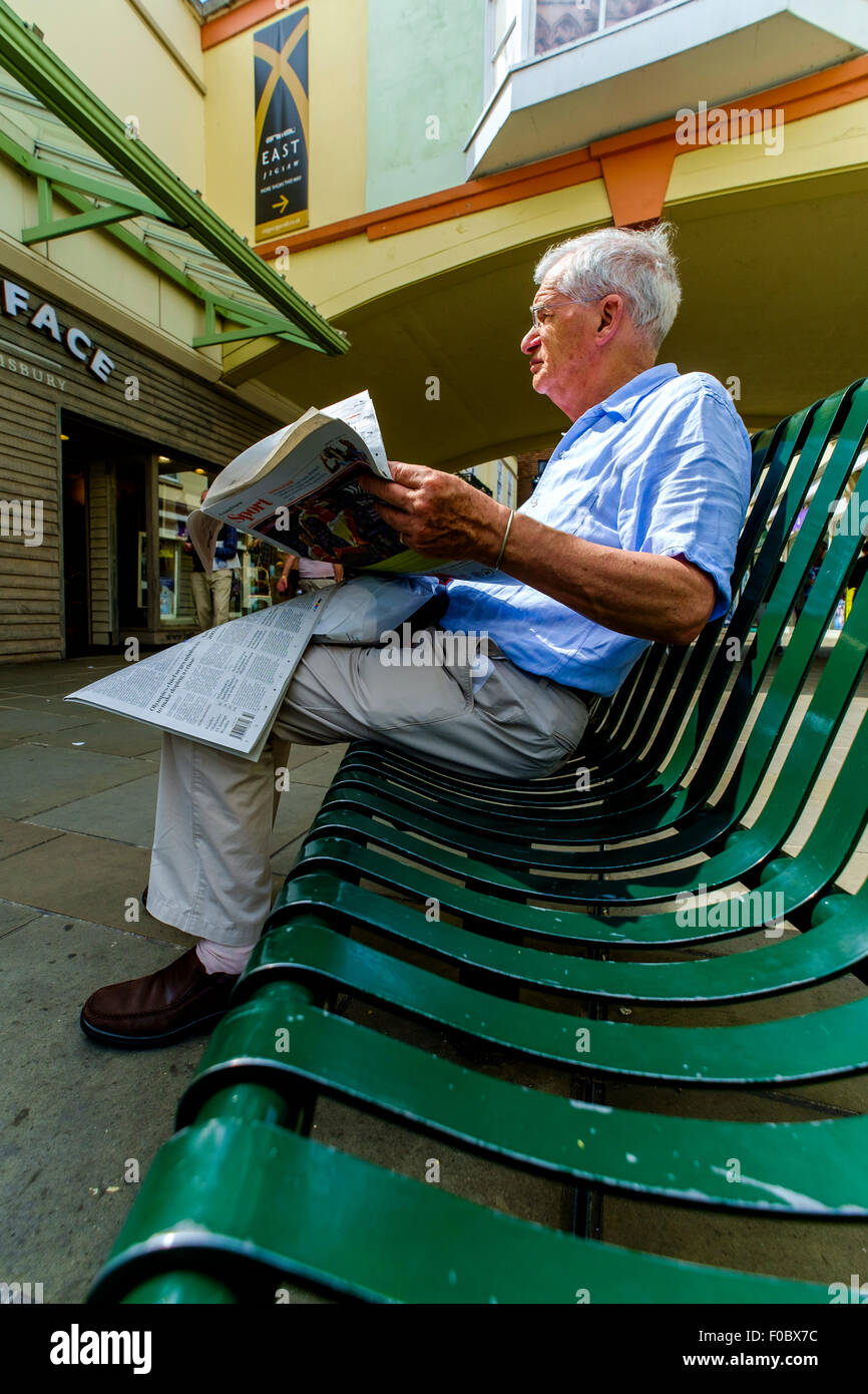 Male reading a newspaper sat on a bench in shopping centre Salisbury ...