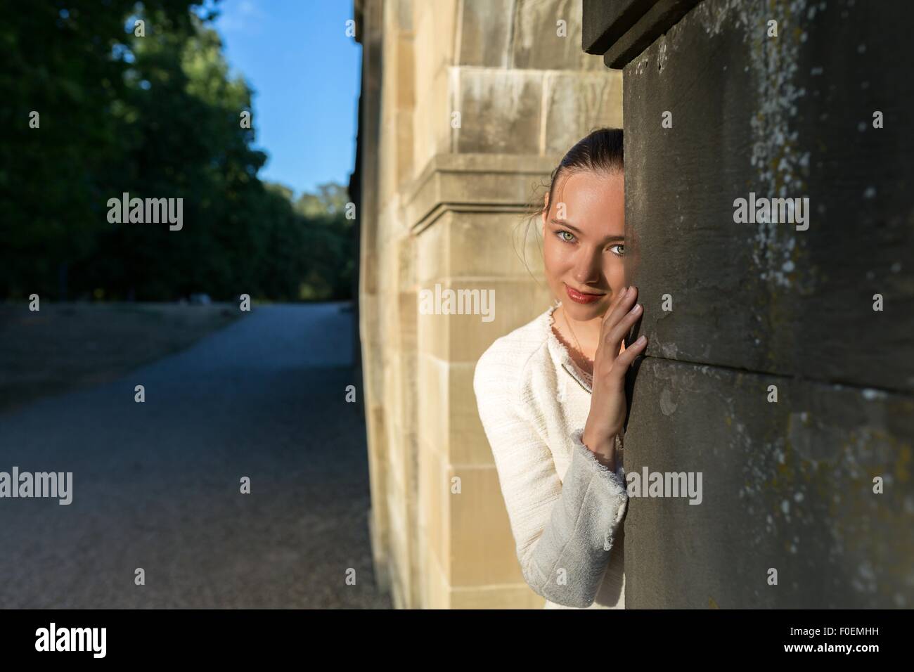 Confident young woman hiding behind a wall and smiling into camera ...