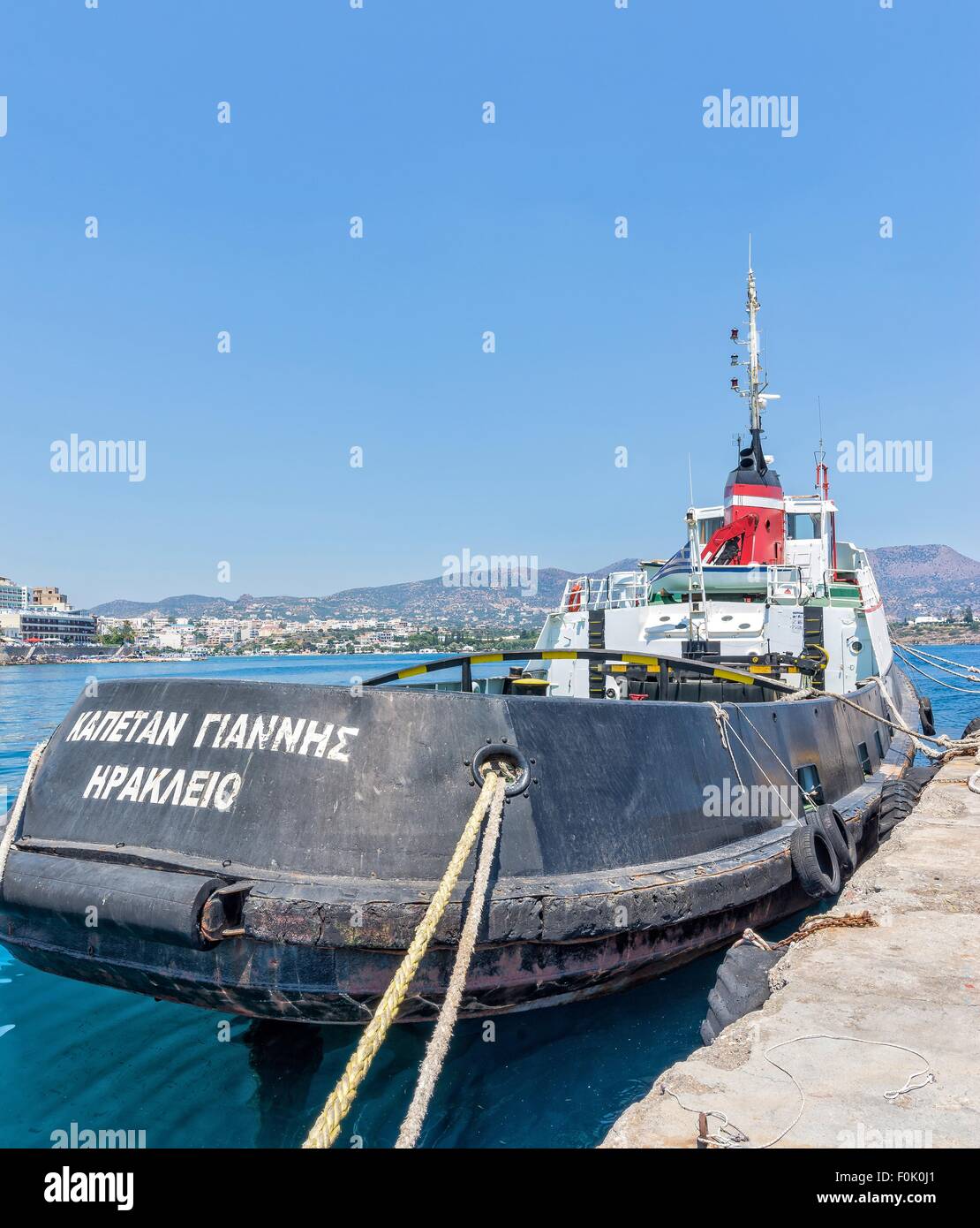 Greek tug boat moored in harbour, with buildings of a town and hillsin ...