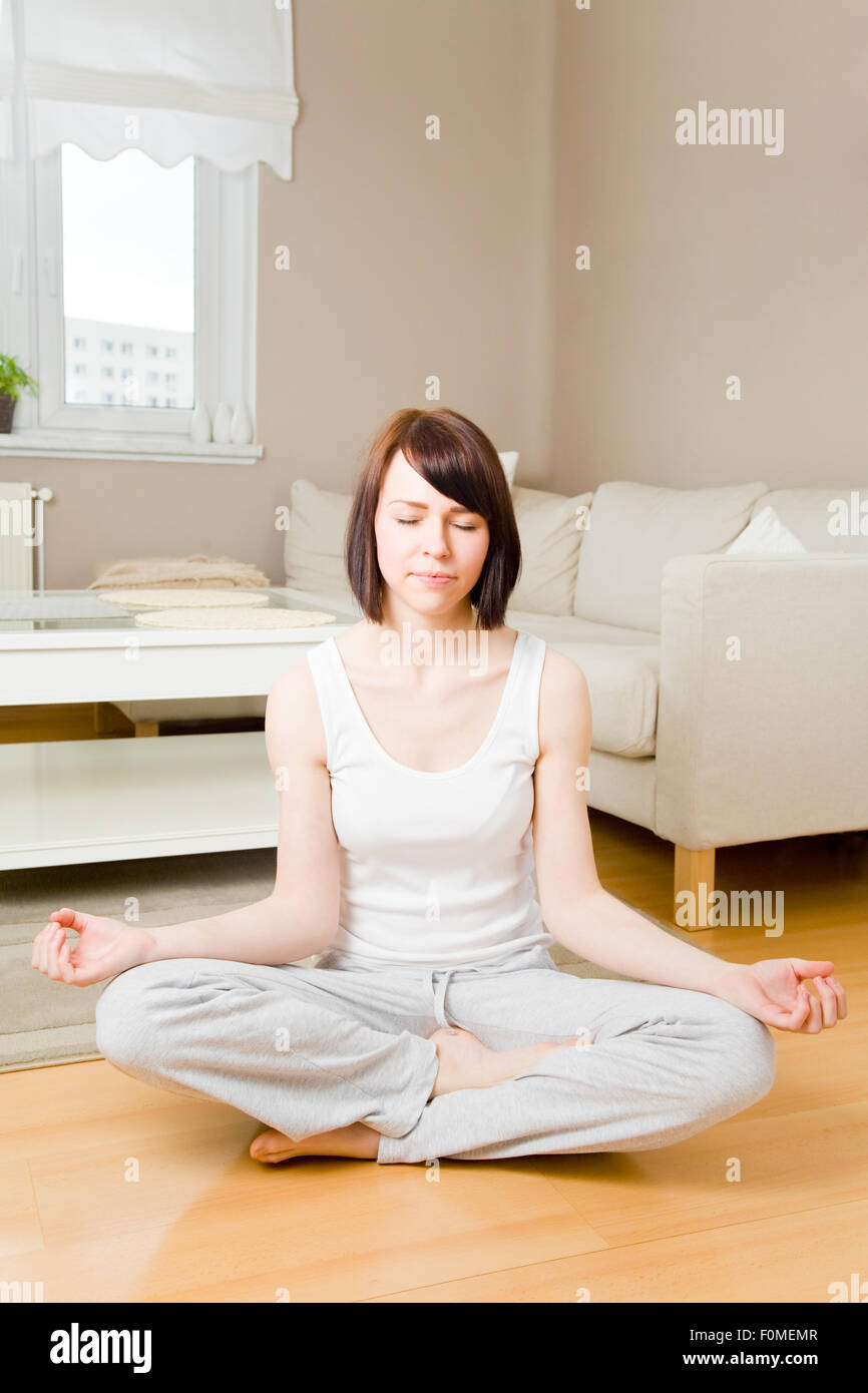 Young woman sitting cross-legged and meditating in her living room ...