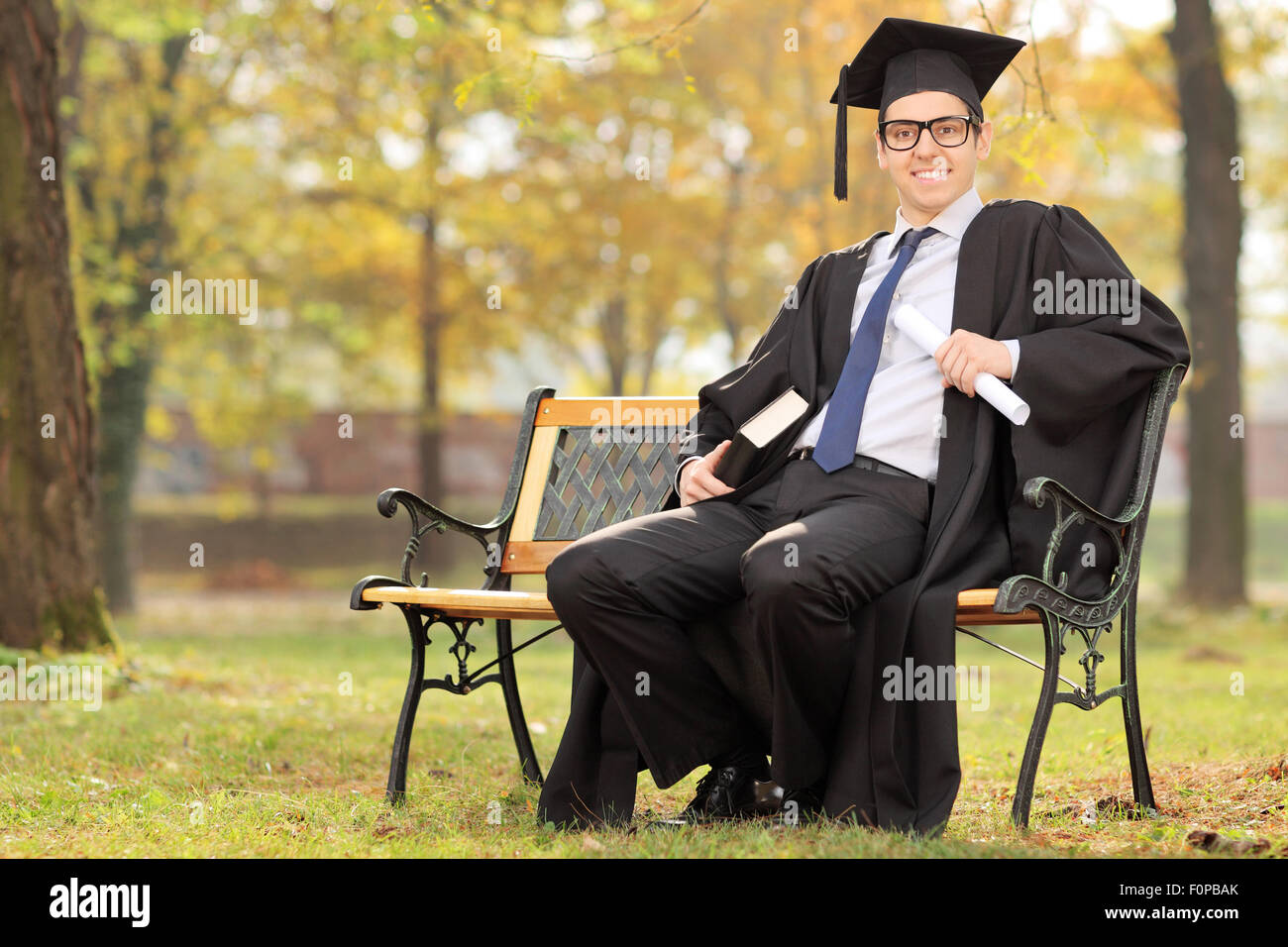 Graduate student holding diploma and a book seated on bench in park ...