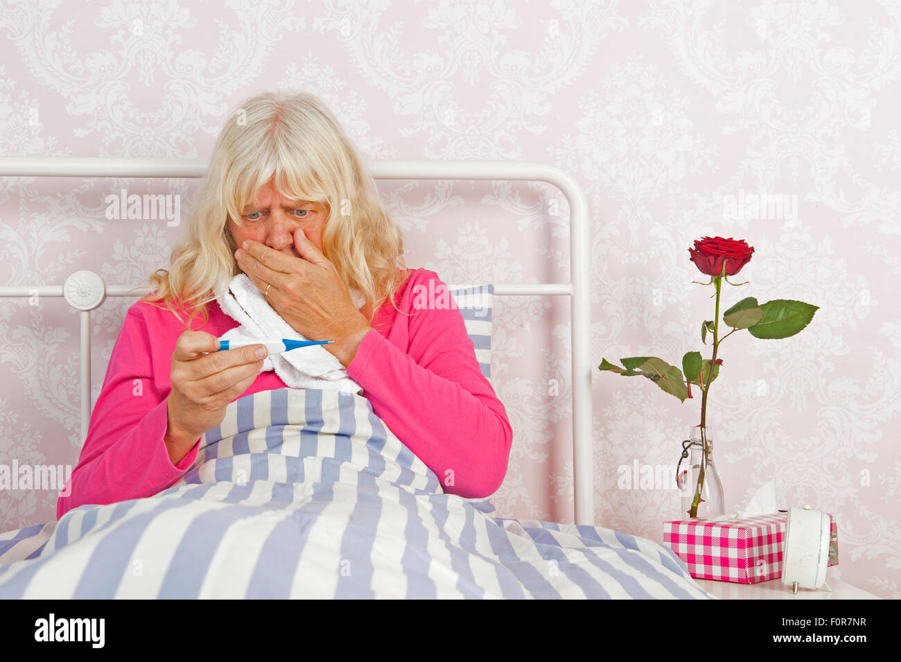 Sick woman sitting in bed looking worried at thermometer with tissues ...