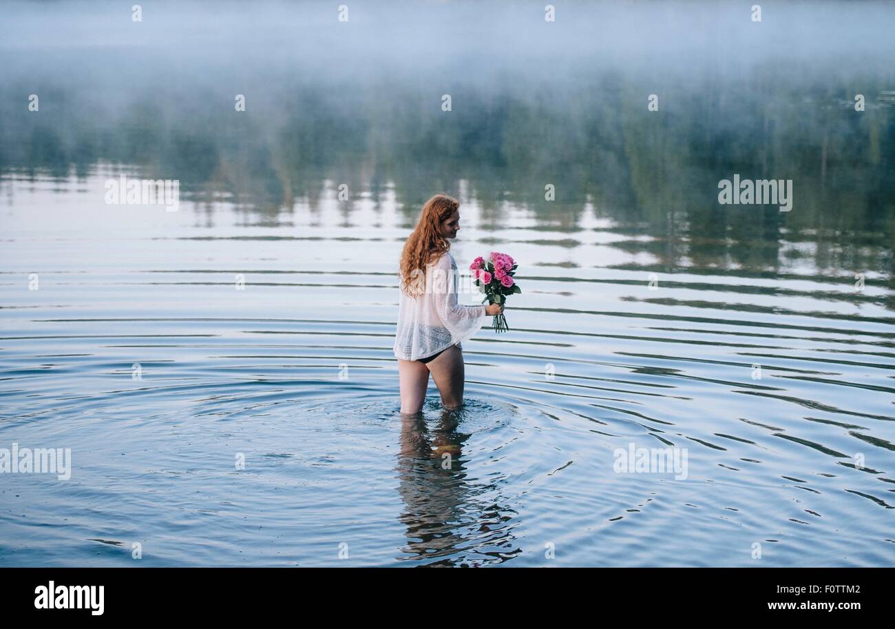 Rear view of young woman wading in misty lake holding bunch of pink ...