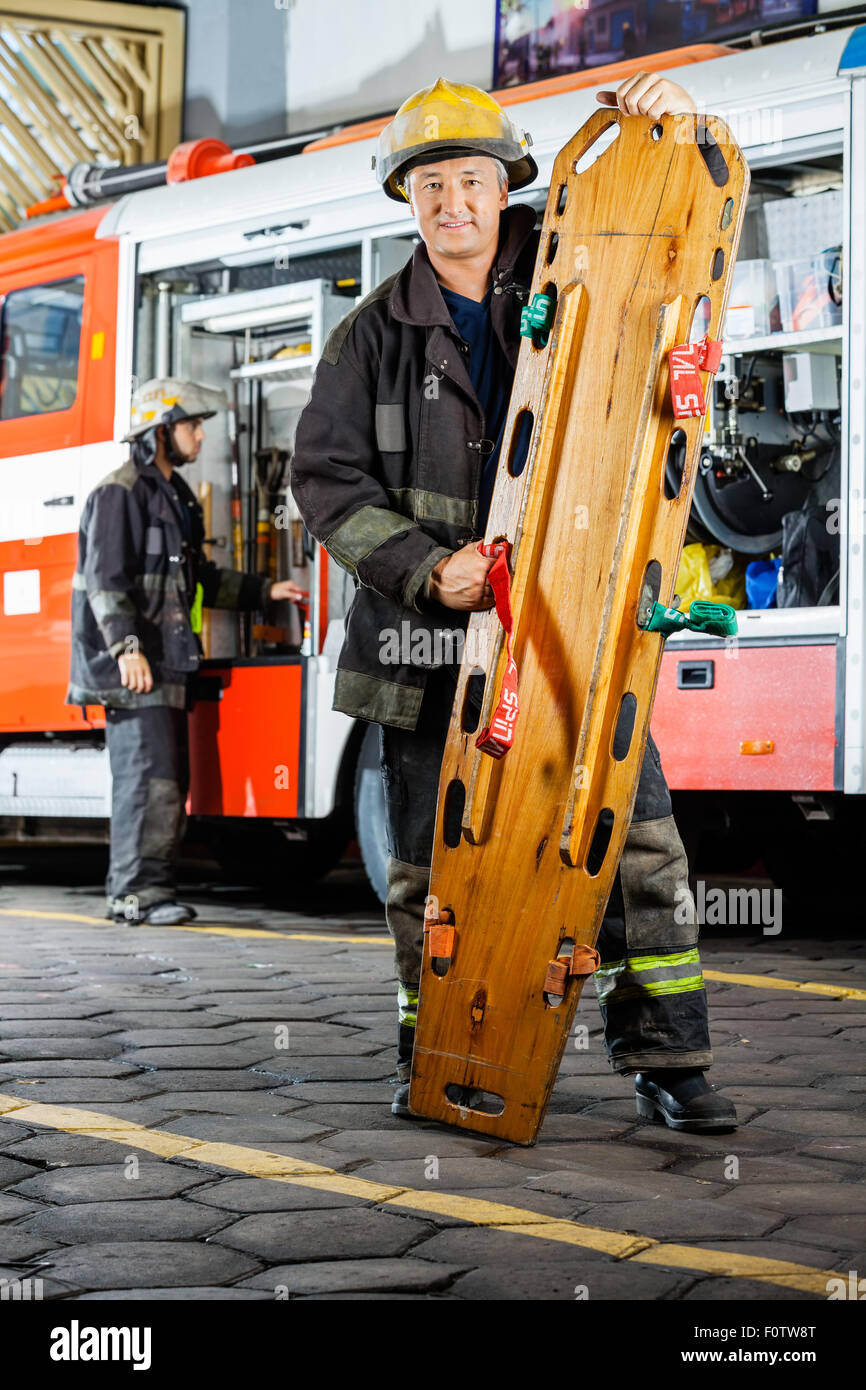 Full length portrait of confident firefighter holding wooden stretcher ...
