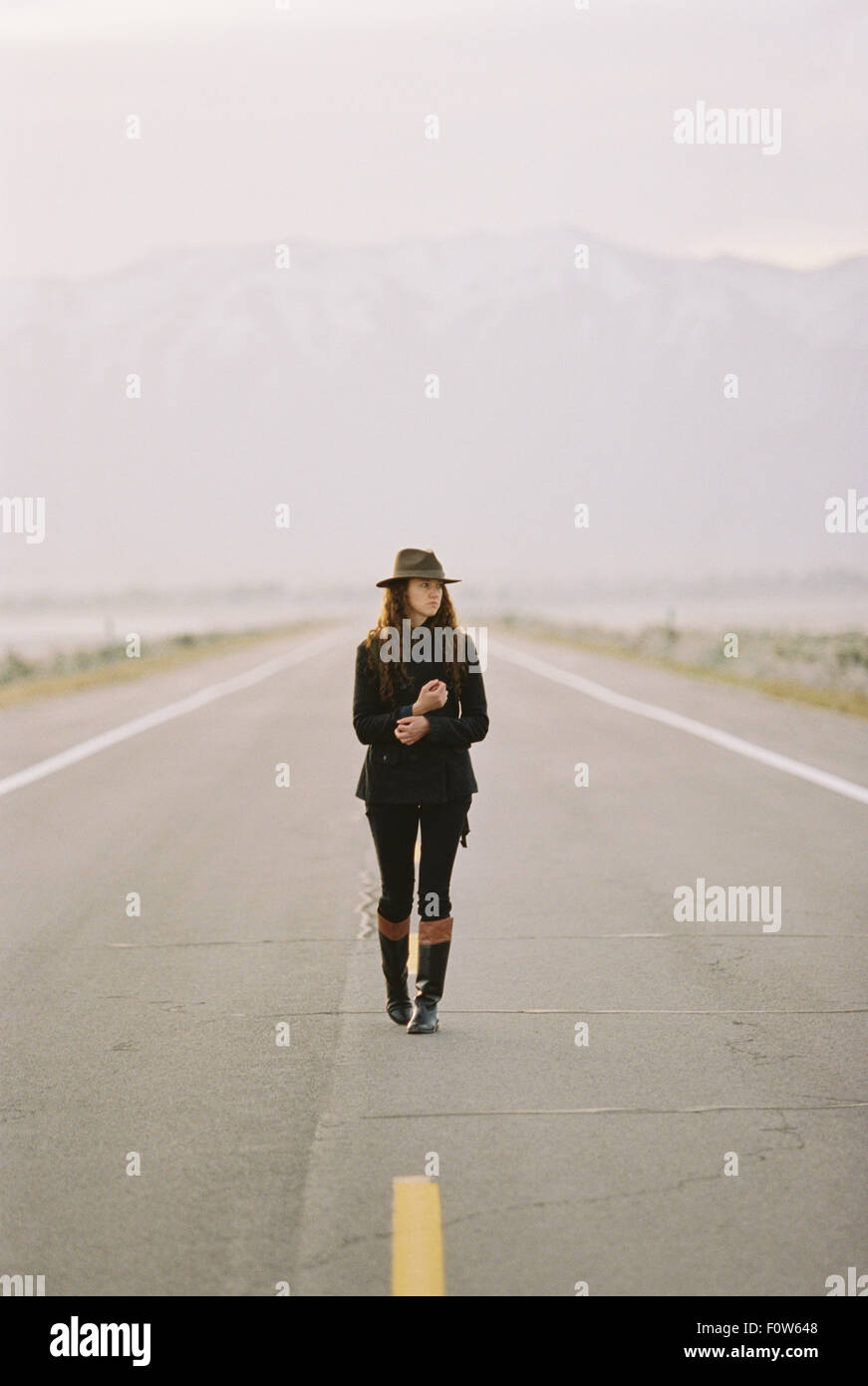 A woman walking down the middle of a country road, wearing a hat and ...