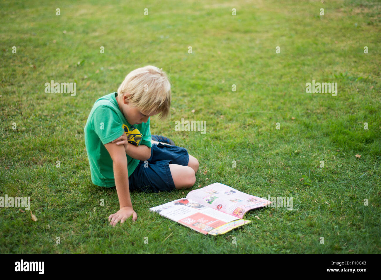 Child blond boy reading magazine in the park, sitting onthe green grass ...