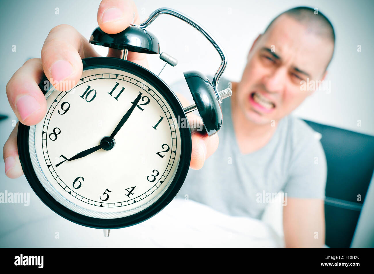 an angry young caucasian man in bed holds in his hand his alarm clock ...