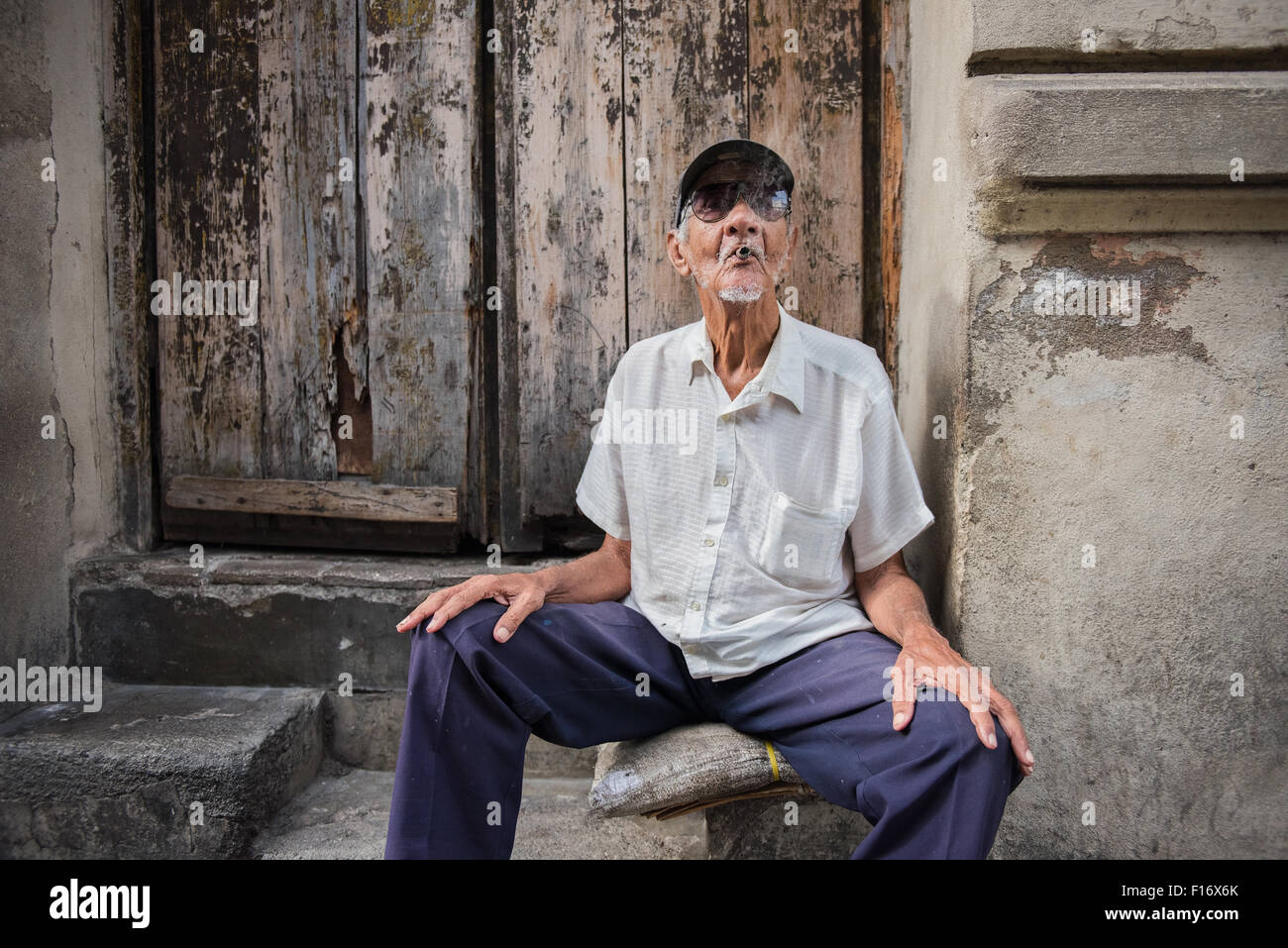 An elderly Cuban man is smoking cigar while sitting by the door of the ...