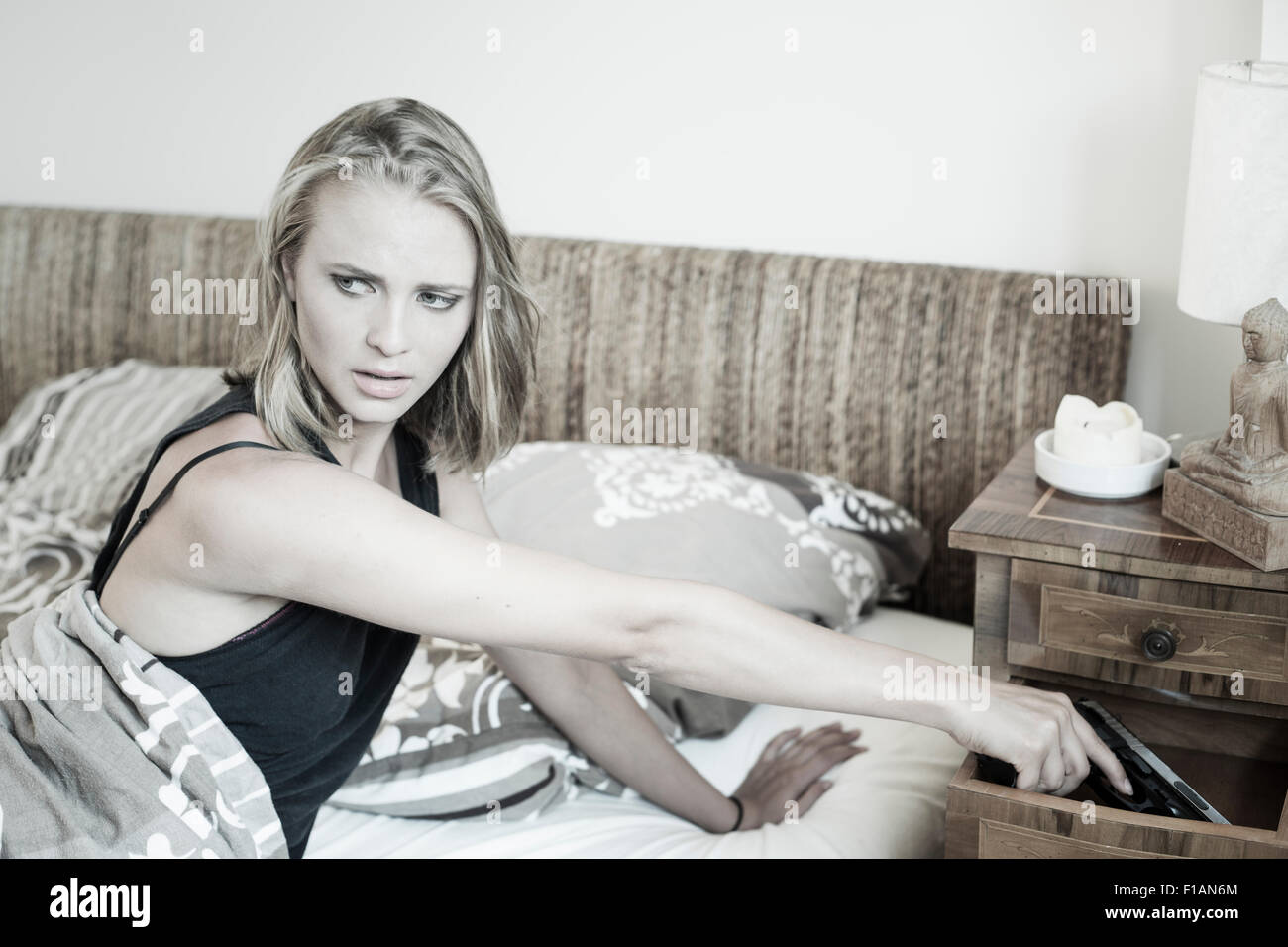 Anxious woman sitting in her bed reaching for pistol in bedside cabinet ...