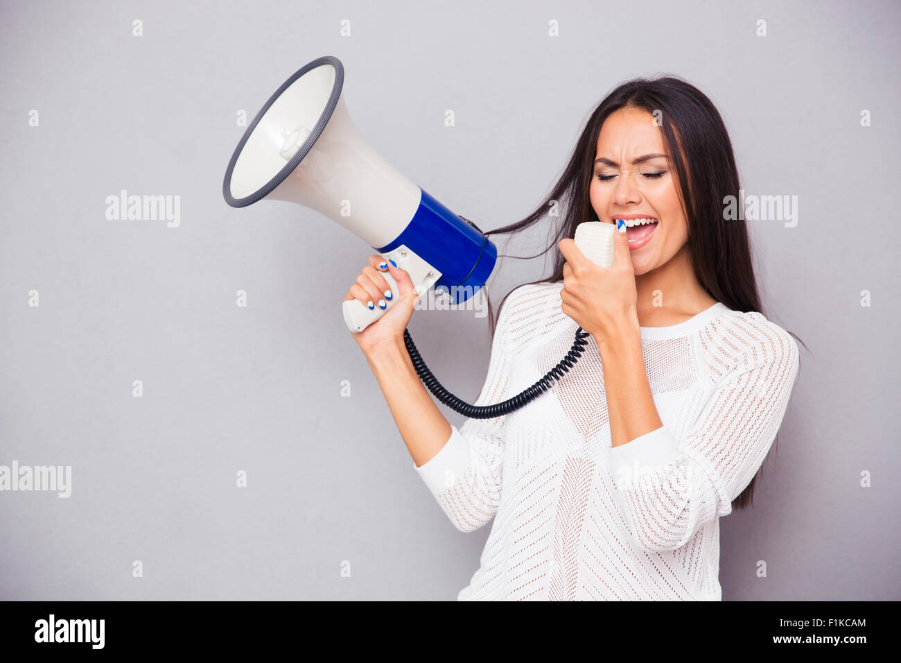 Portrait of a casual beautiful woman screaming on megaphone over gray ...