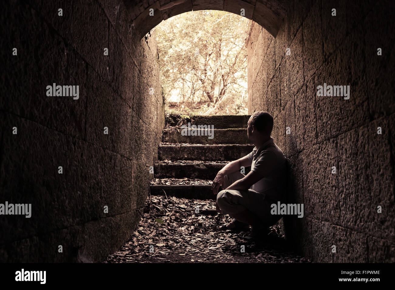 Young man sits inside of dark stone tunnel with glowing end, warm tonal ...