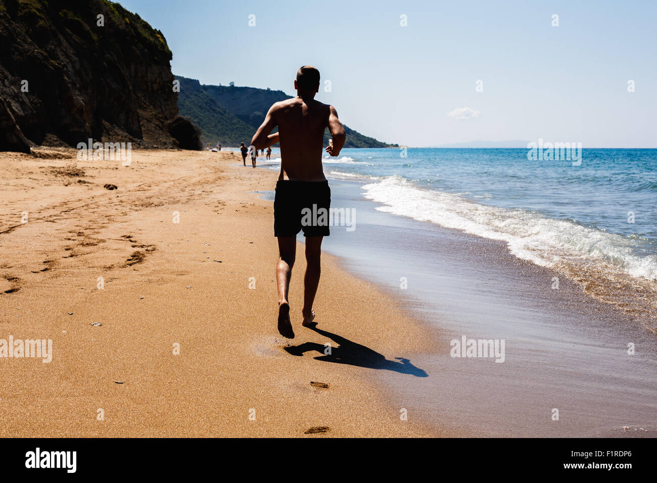 Back view of a runner man running on the beach at the middle of a day ...
