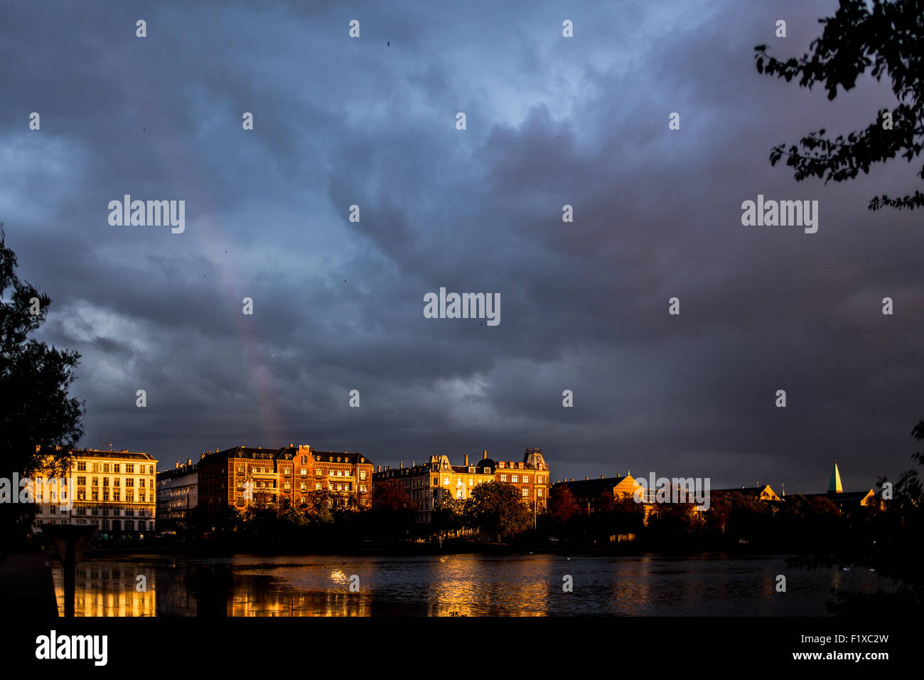 Sun hits buildings with dark skies and a rainbow behind Stock Photo - Alamy