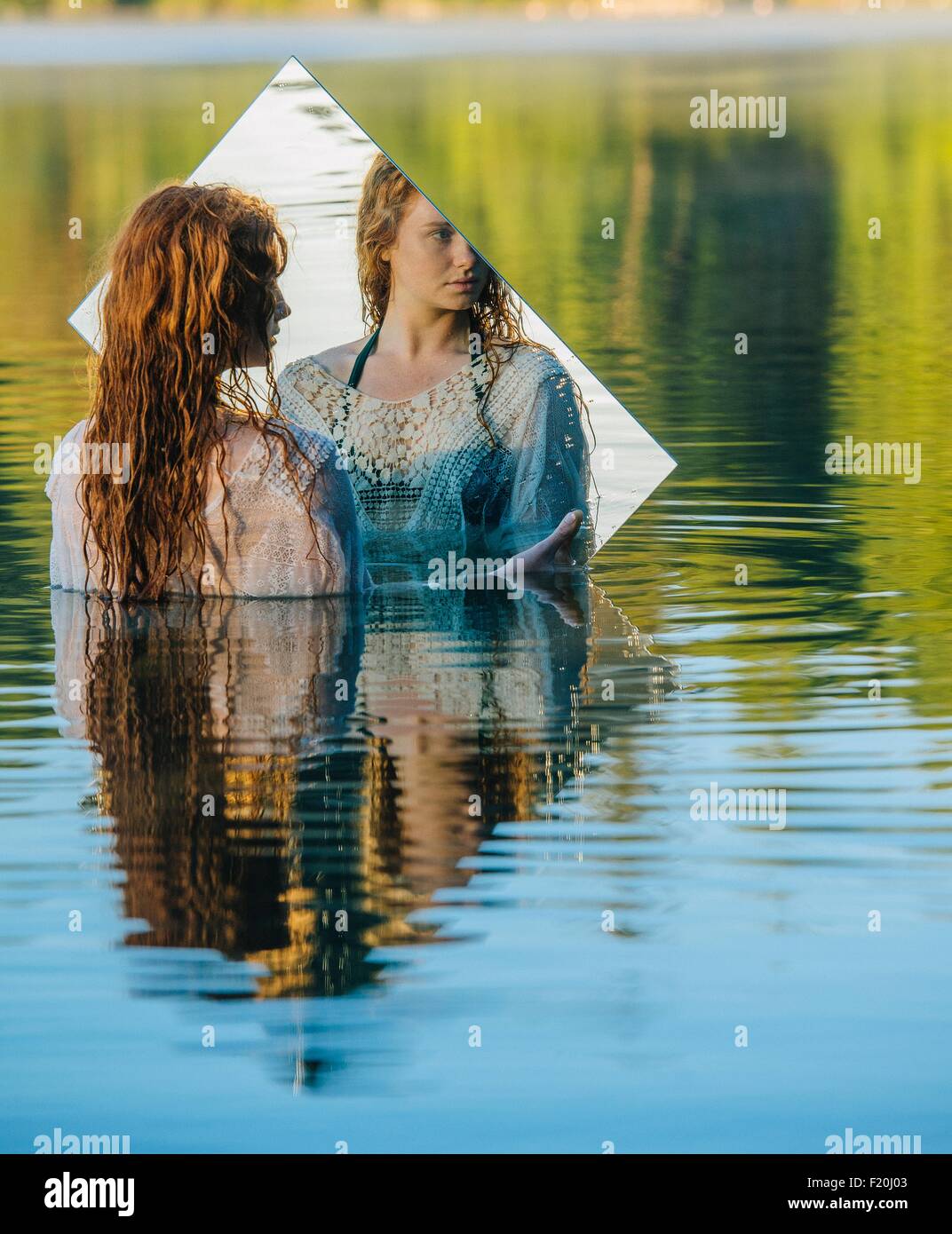Young woman standing in lake, holding mirror, looking at reflection ...