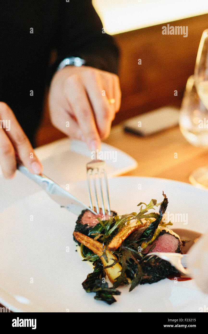 A man seated using a knife and fork eating meat and roasted vegetables ...