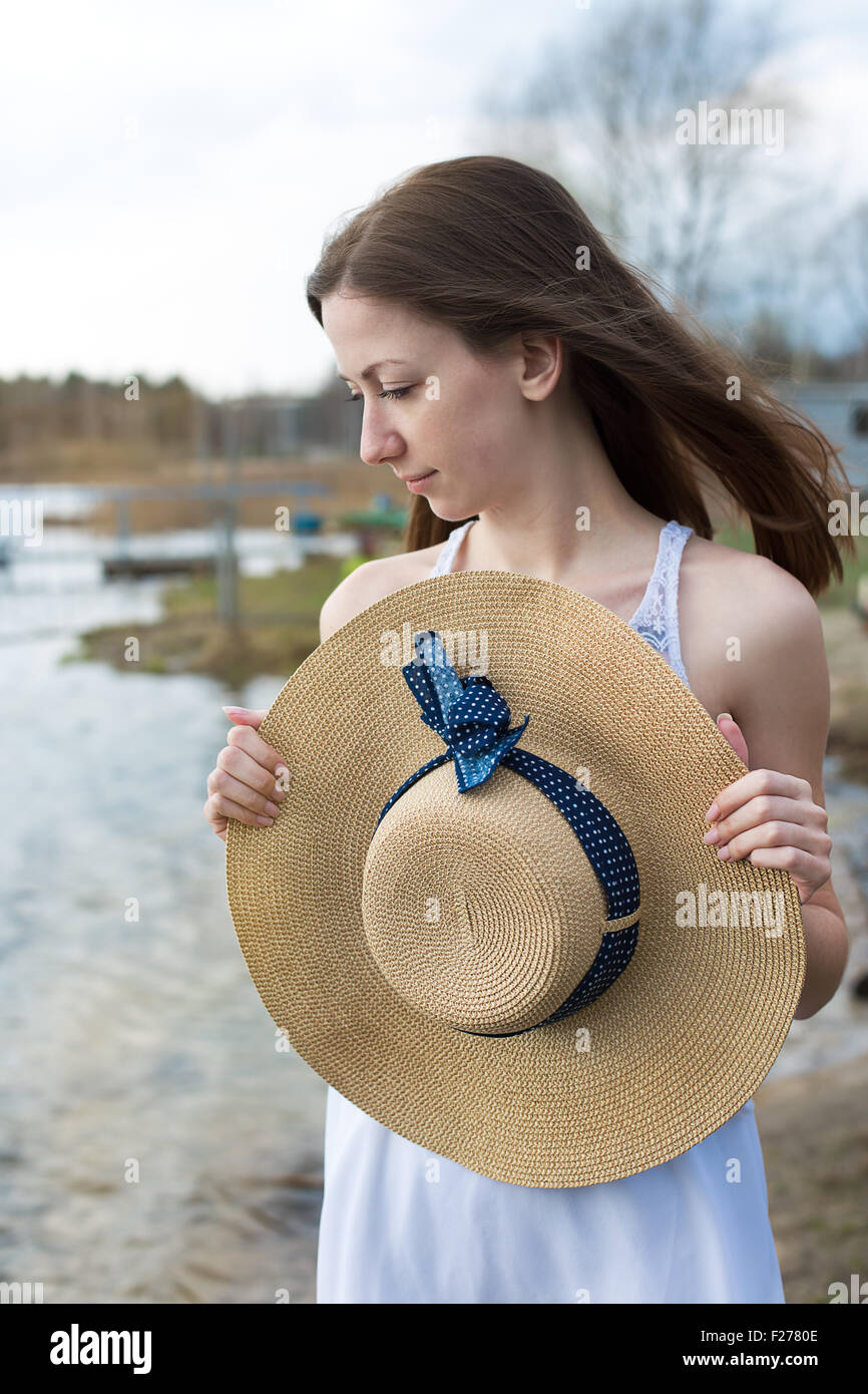 Freckled happy girl in white dress holding thatch hat in his hands and looking to the side in ...