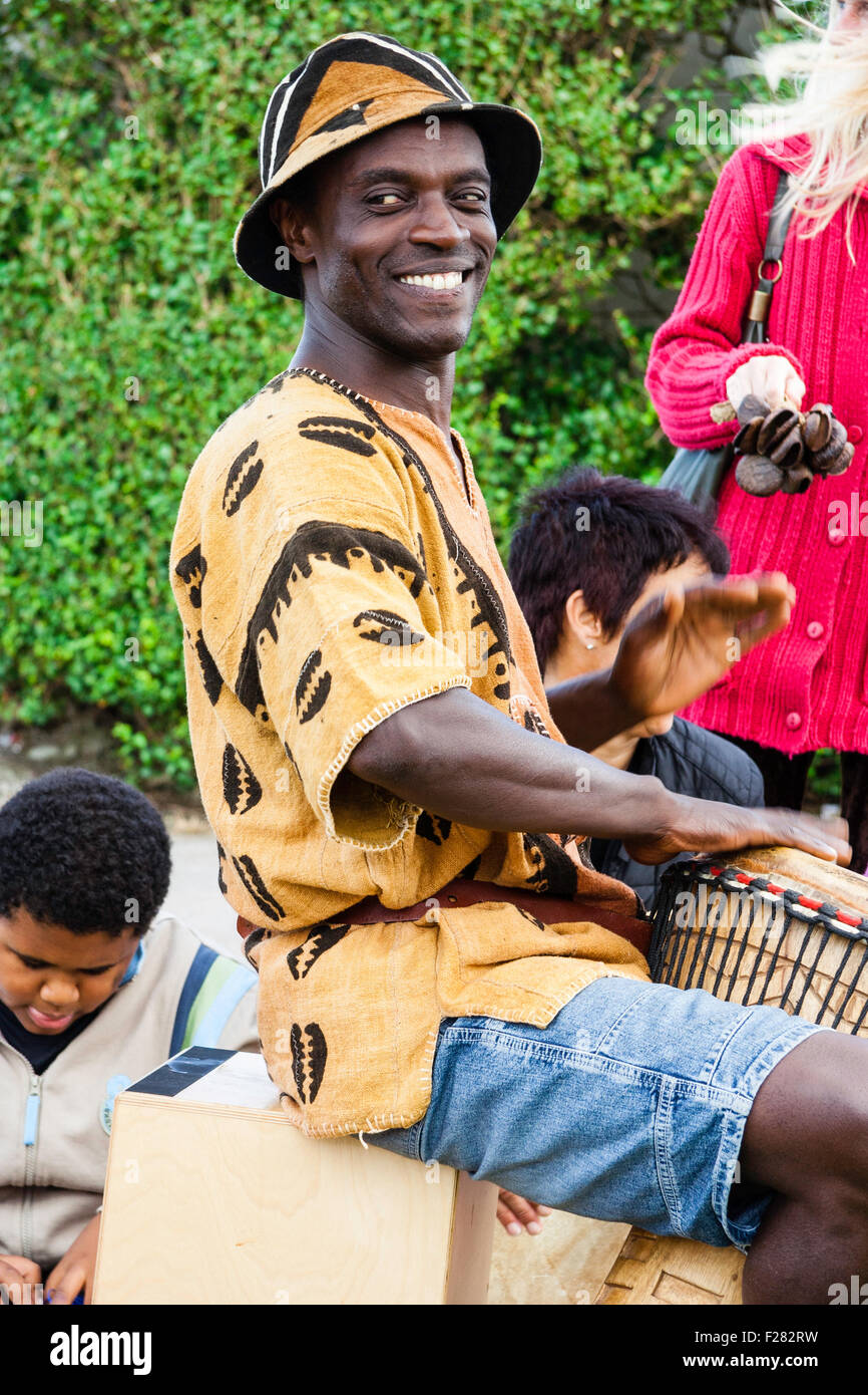 Black African man in shorts, shirt and hat sitting playing bongo drum ...