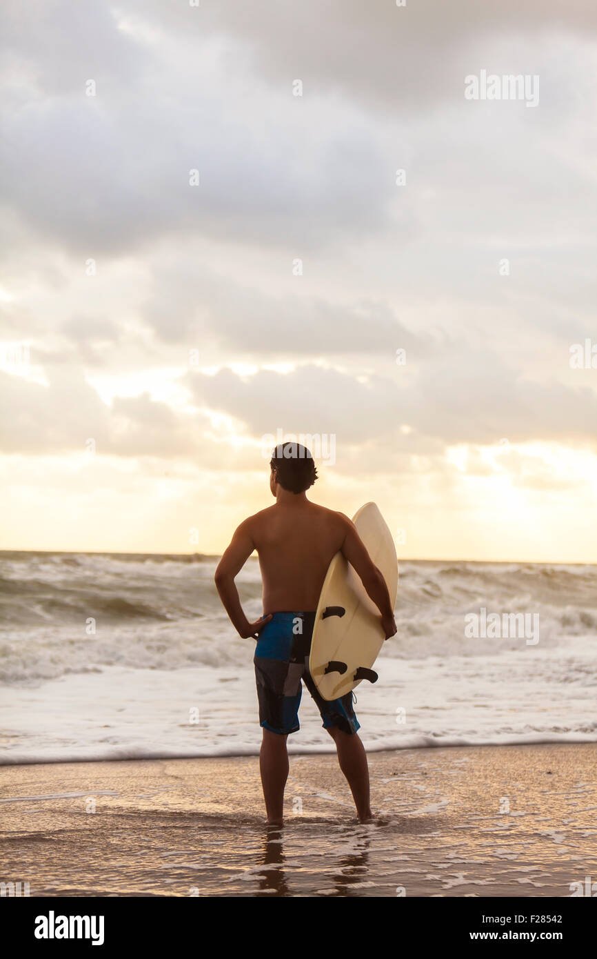 Rear view of young man surfer with white surfboard on a beach at sunset ...
