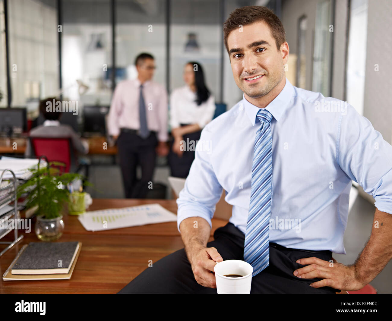 portrait of caucasian business executive sitting on desk in office ...
