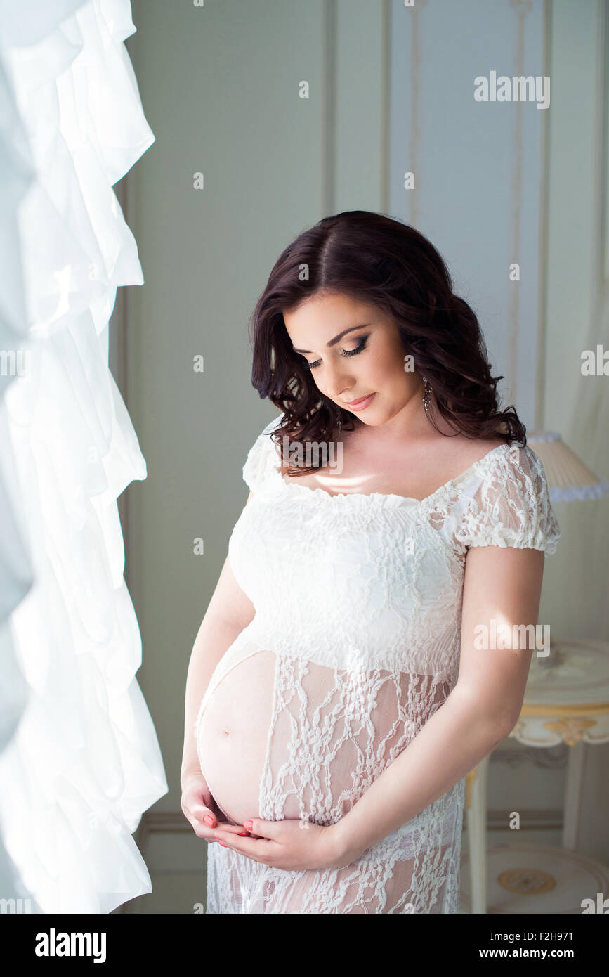 Beautiful pregnant mom in white lace dress stand at window and look on ...