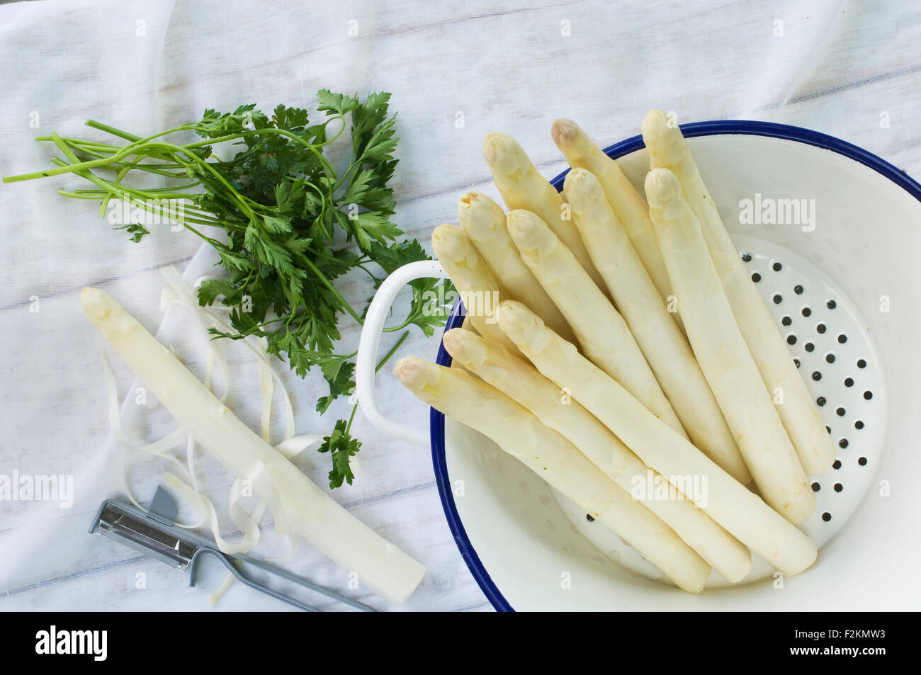 Colander of white asparagus, flat leaf parsley and peeler on white ...