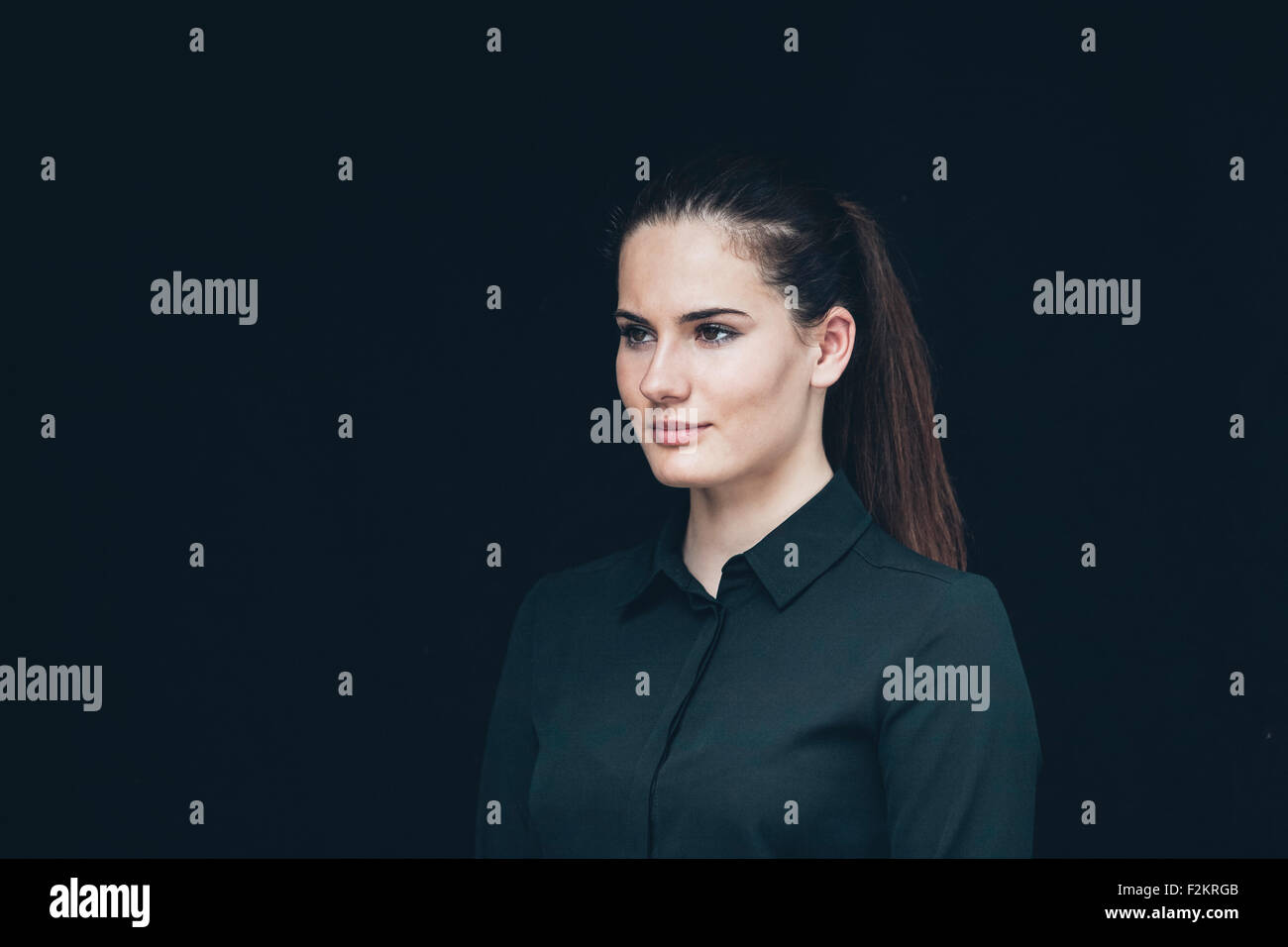 Portrait of young woman with ponytail in front of black background ...