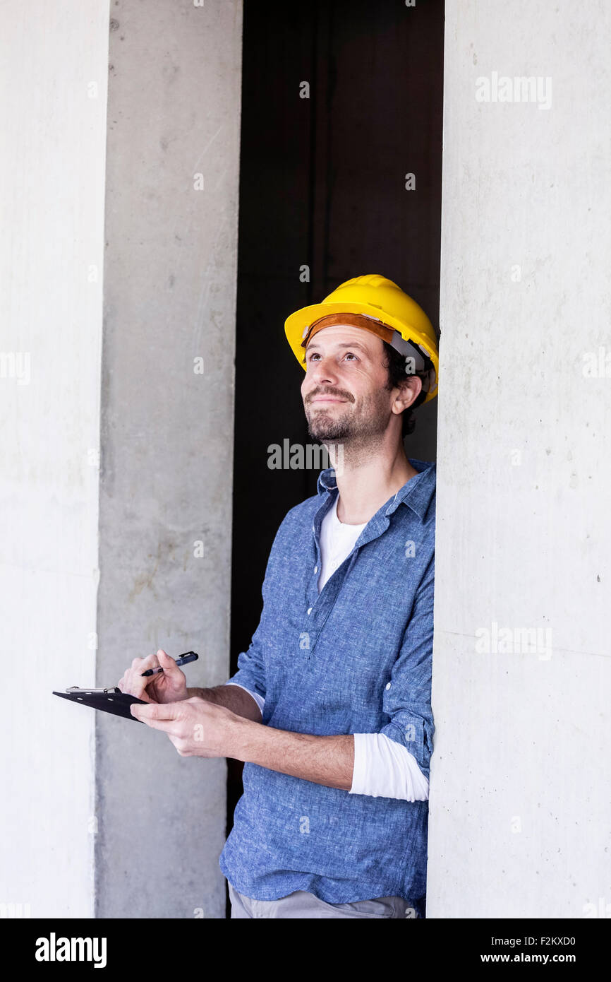Man with hard hat on construction site looking up writing on clipboard ...