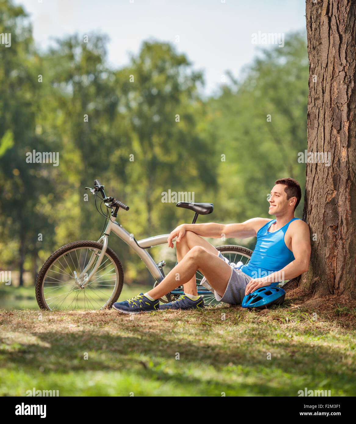 Young male cyclist resting by a tree in park with his bicycle beside ...